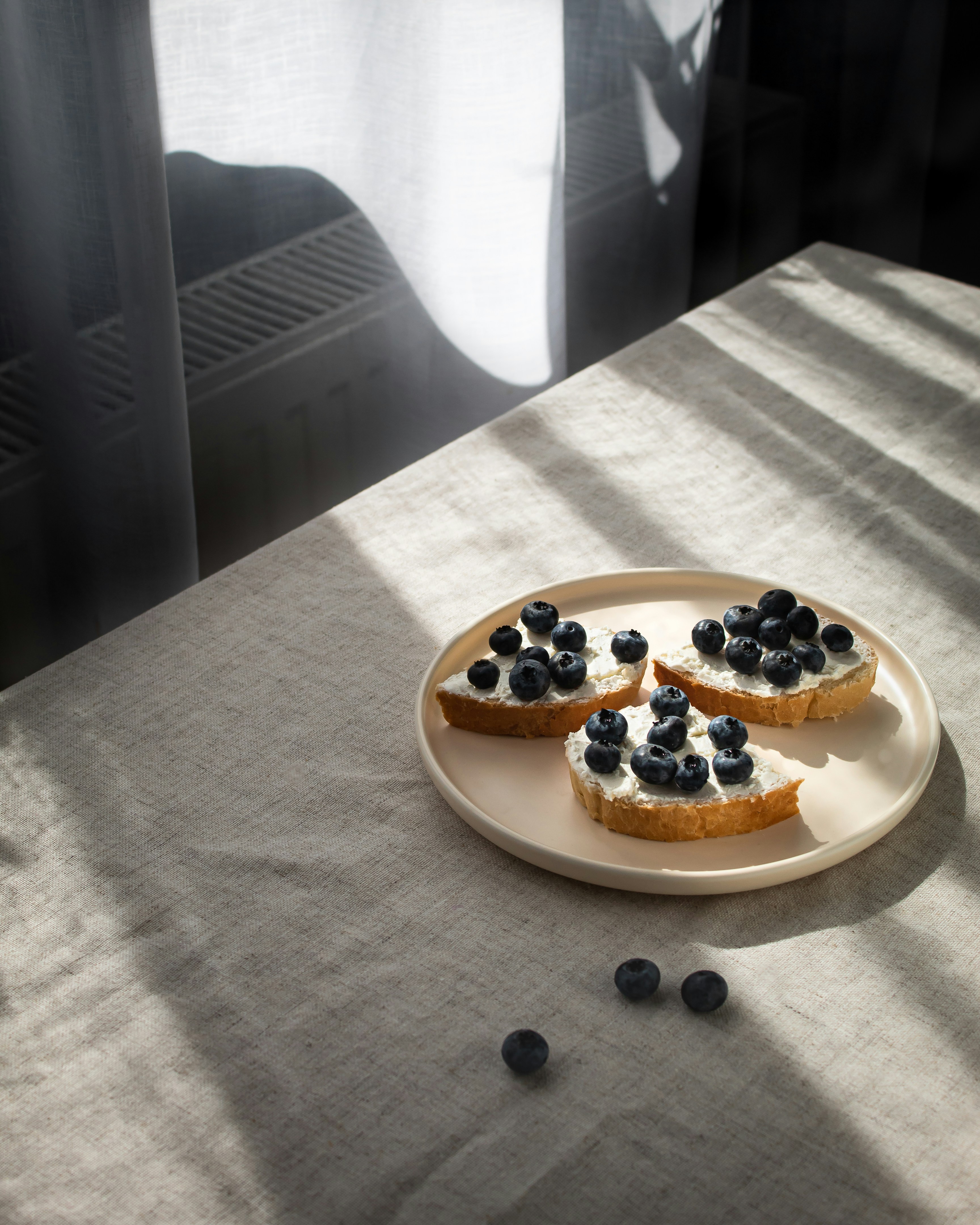 Three blueberry-topped toast slices rest on a pale plate, bathed in soft window light with a few berries scattered on the linen table.
