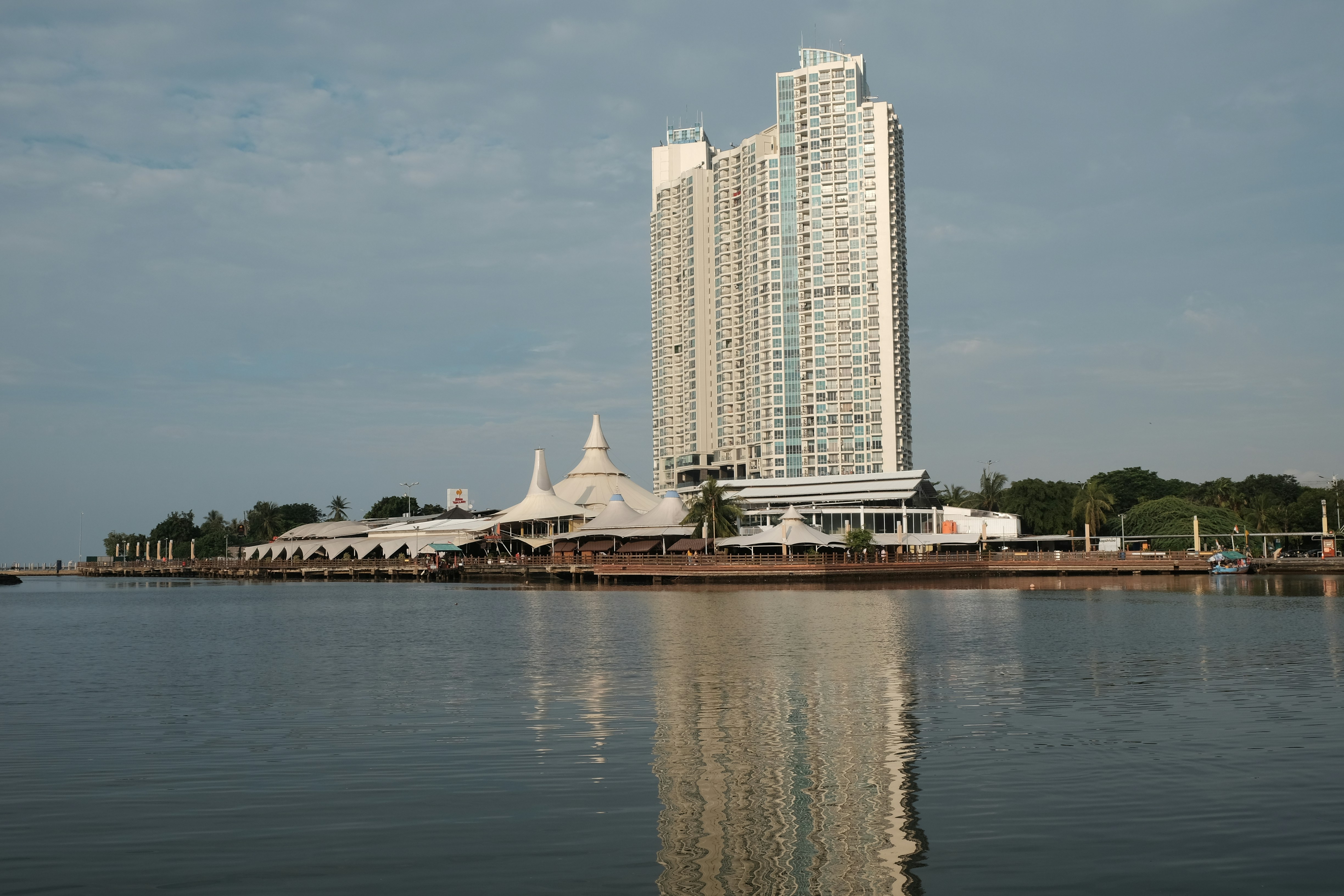 High-rise building and modern waterfront structure reflected in calm waters under a cloudy sky.