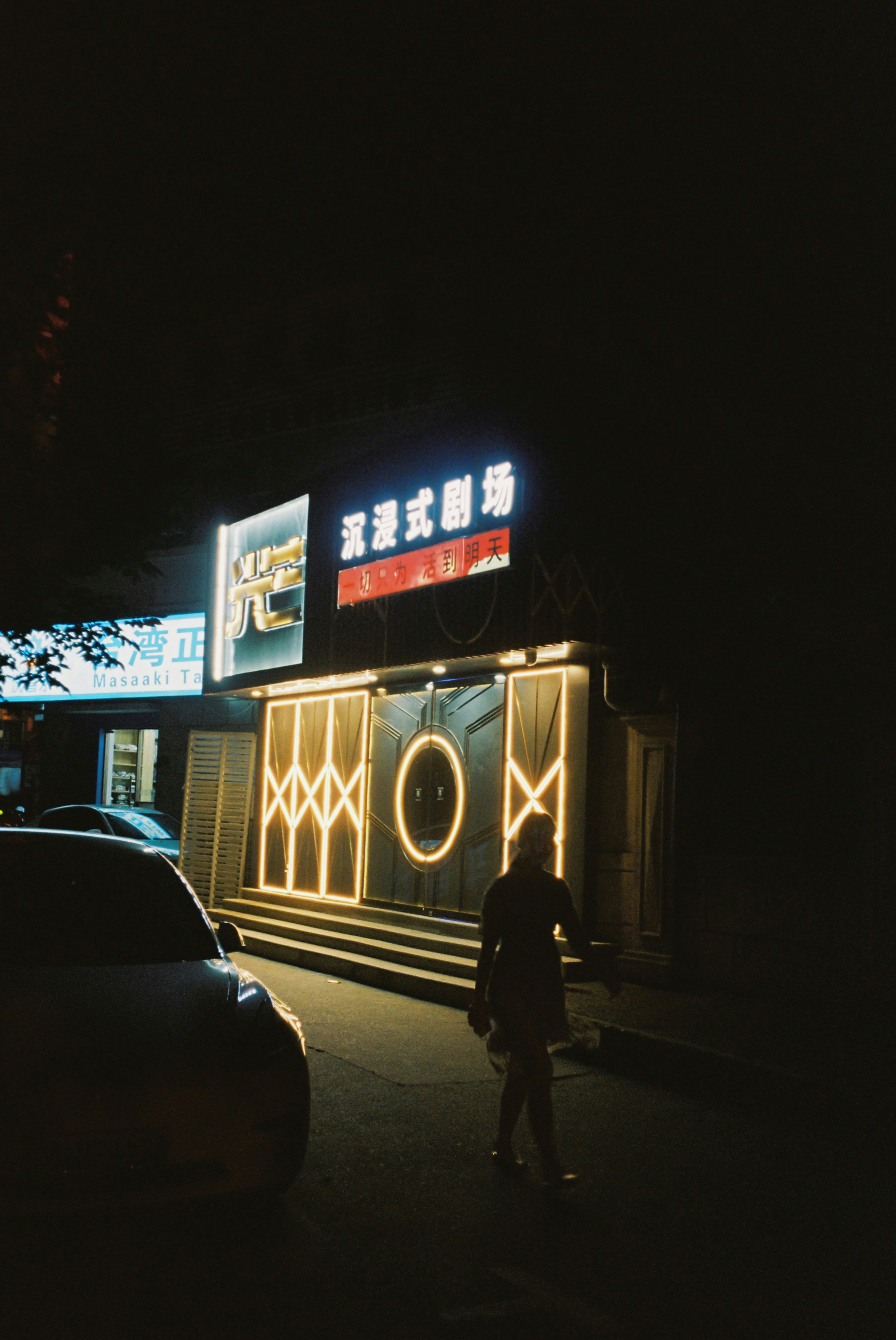 Vibrant neon signage illuminates a theater entrance at night, casting a warm glow on the surrounding street. A figure walks by, adding a sense of life to the scene.