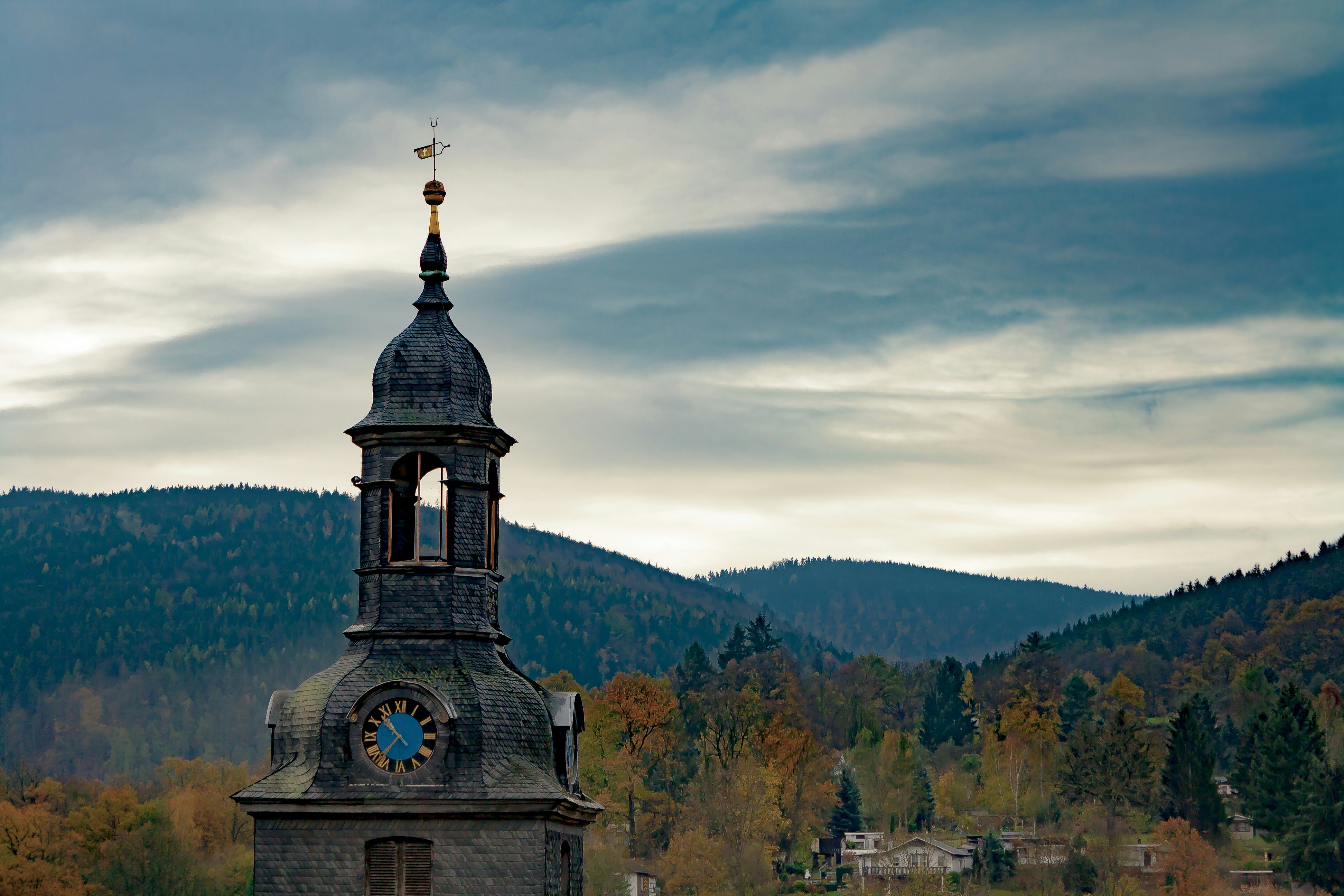 Clock tower rises above autumn foliage, framed by rolling hills under a moody sky.