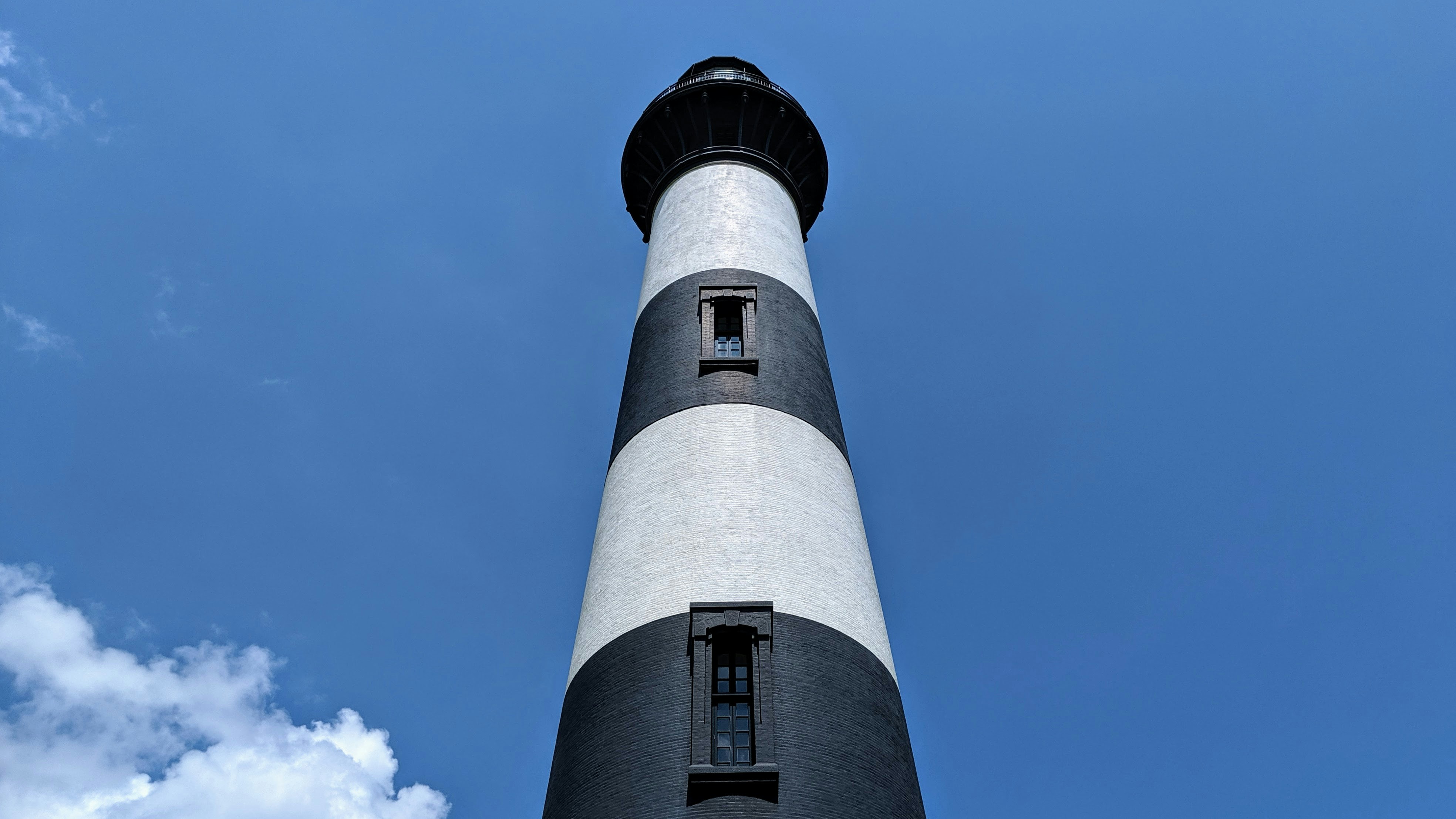 A tall lighthouse with bold black and white stripes stands against a clear blue sky, symbolizing maritime guidance. Clouds drift softly at its base.