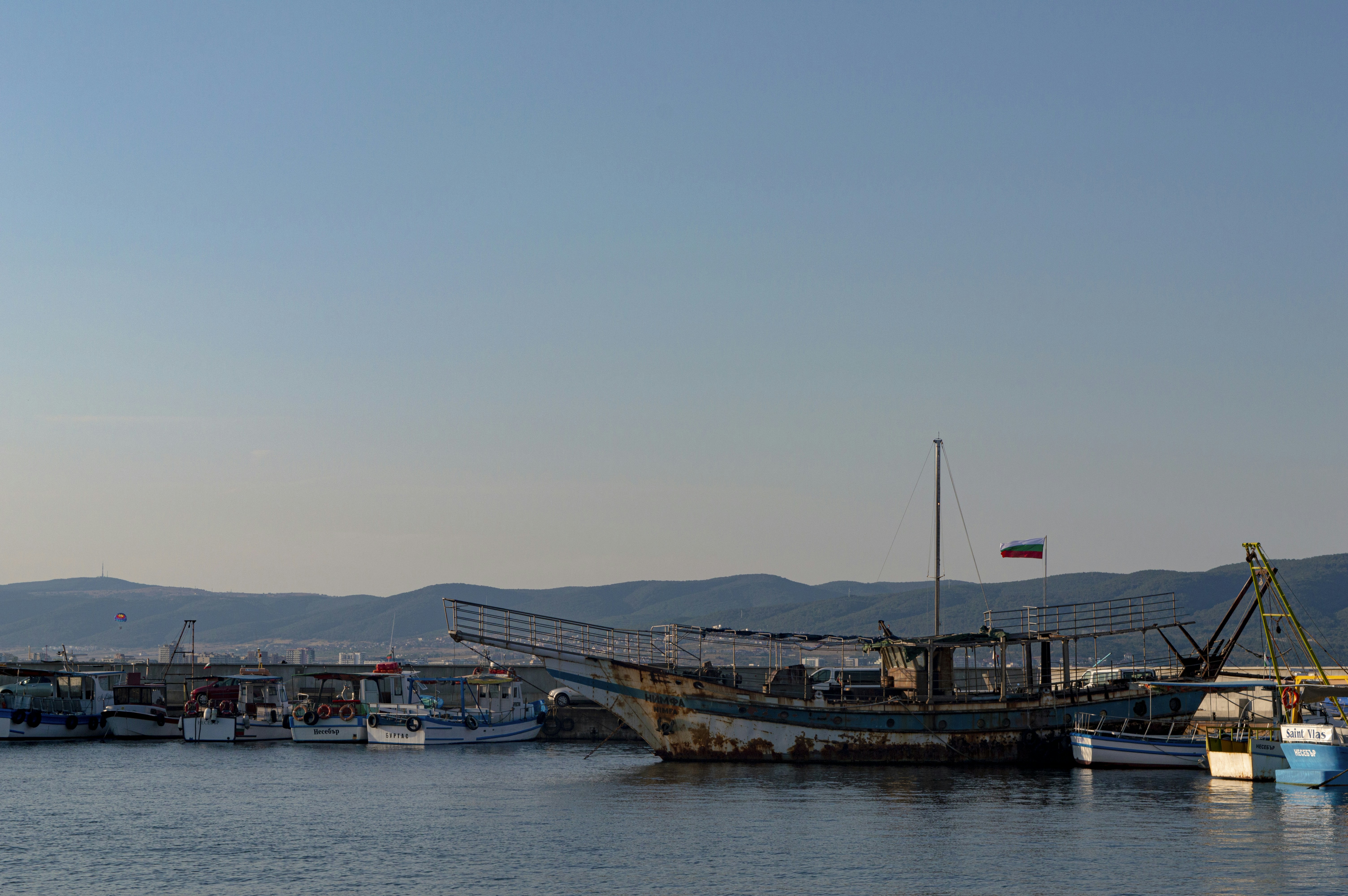 Rustic fishing boat anchored in a tranquil harbor, surrounded by smaller vessels and distant hills under a clear sky.
