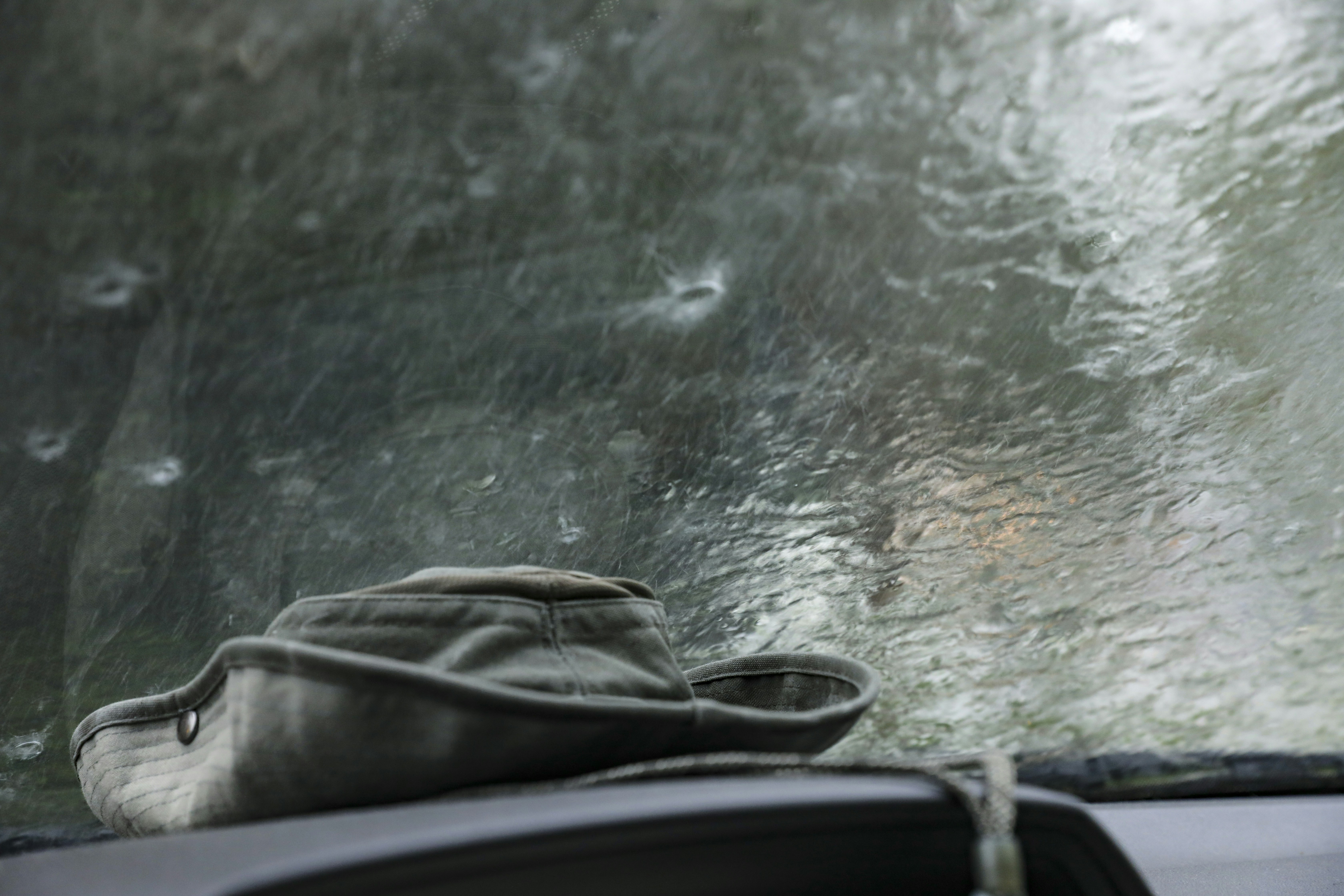 A weathered hat resting on a car dashboard, with rain-soaked glass distorting the view outside. The droplets create a textured backdrop.
