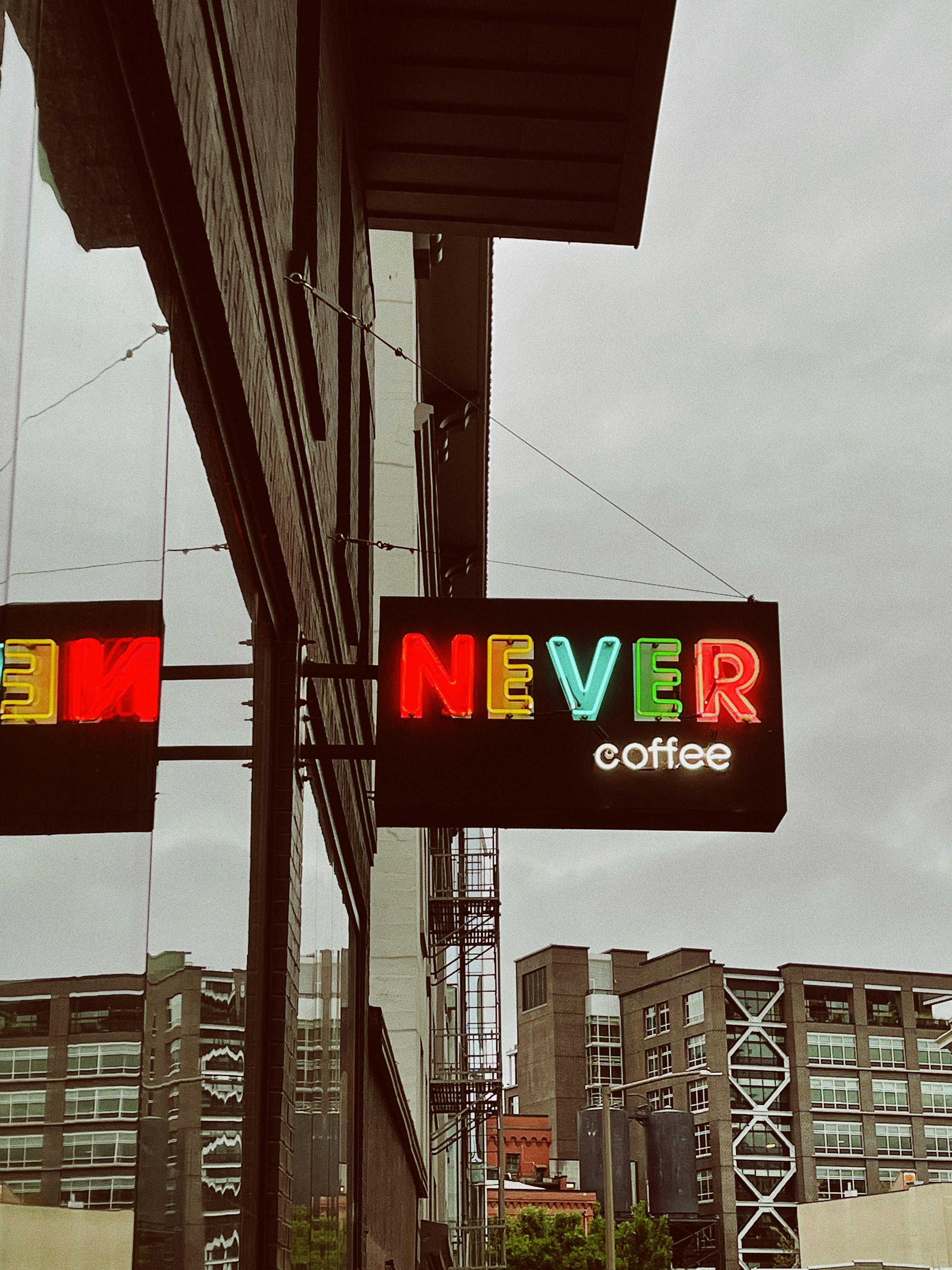 Neon sign reading 'NEVER coffee' hangs outside a modern building, reflecting the urban environment. The cloudy sky adds a moody backdrop.