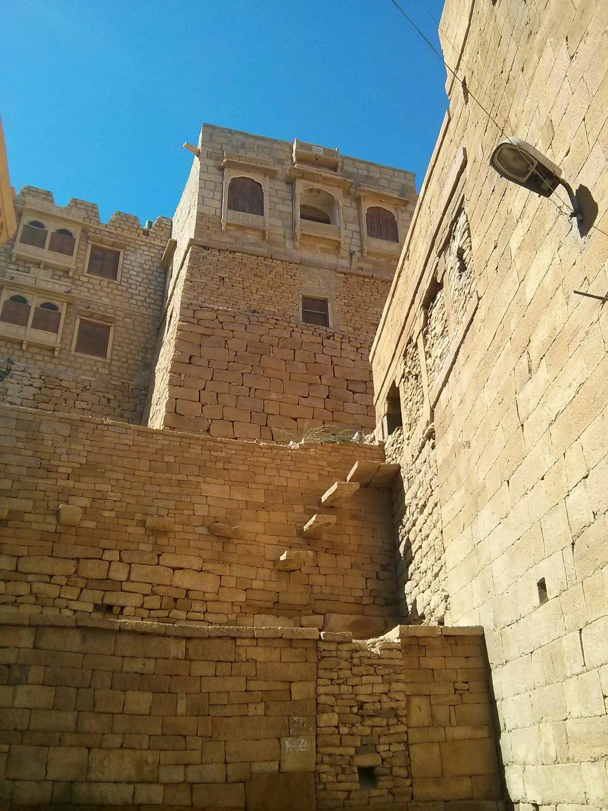 Stone fortress built from layered limestone rises against a bright blue sky. A streetlamp extends from the right wall, anchoring the composition.
