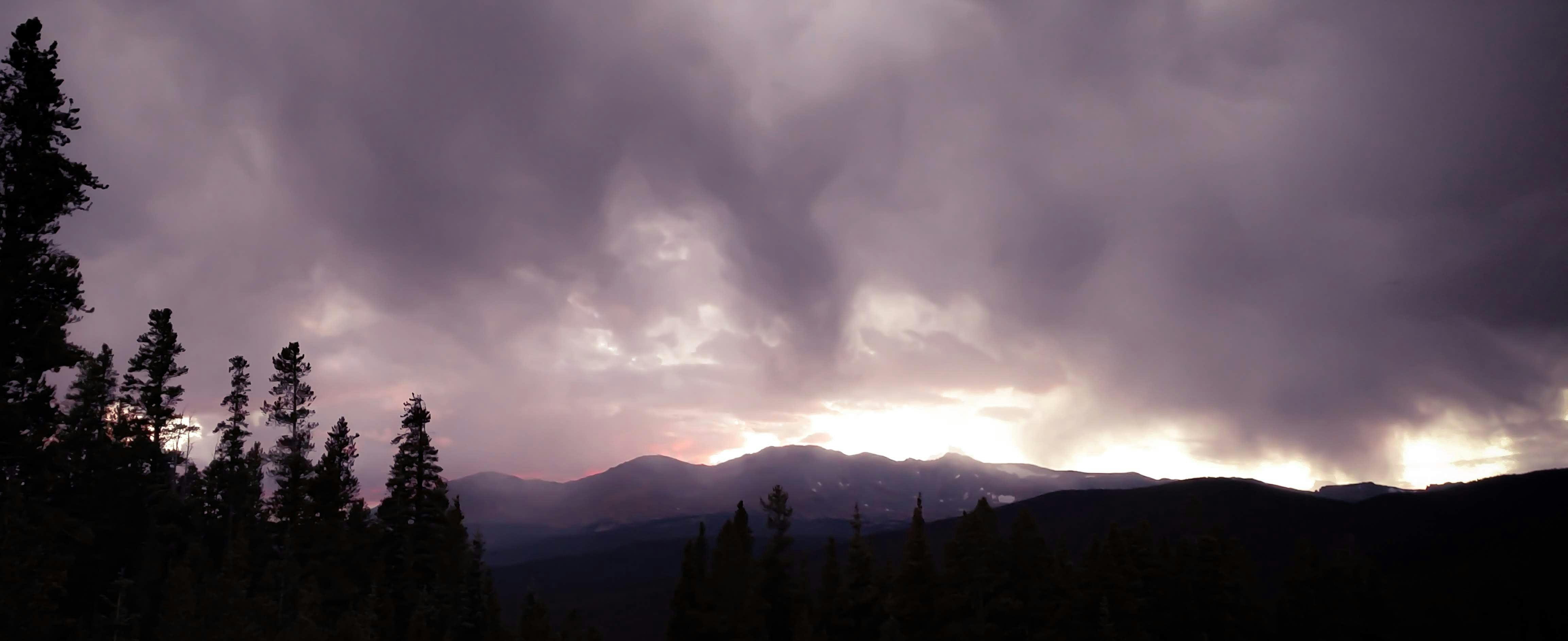 Dramatic clouds loom over a distant mountain range, silhouetted by the fading light. Tall evergreen trees frame the scene, enhancing the sense of depth.