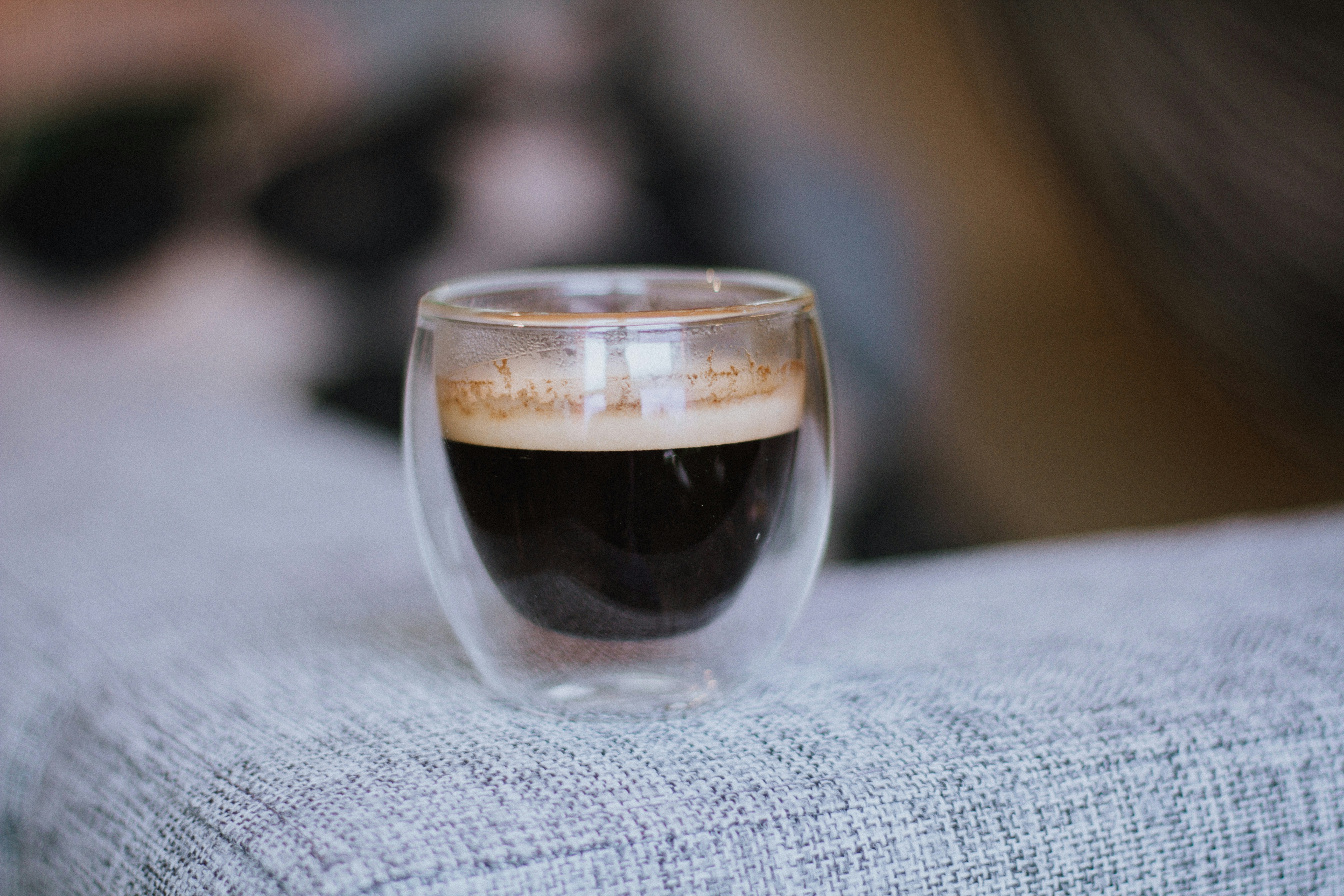 A clear glass cup filled with rich espresso sits on a textured surface, highlighting its creamy froth and deep color.