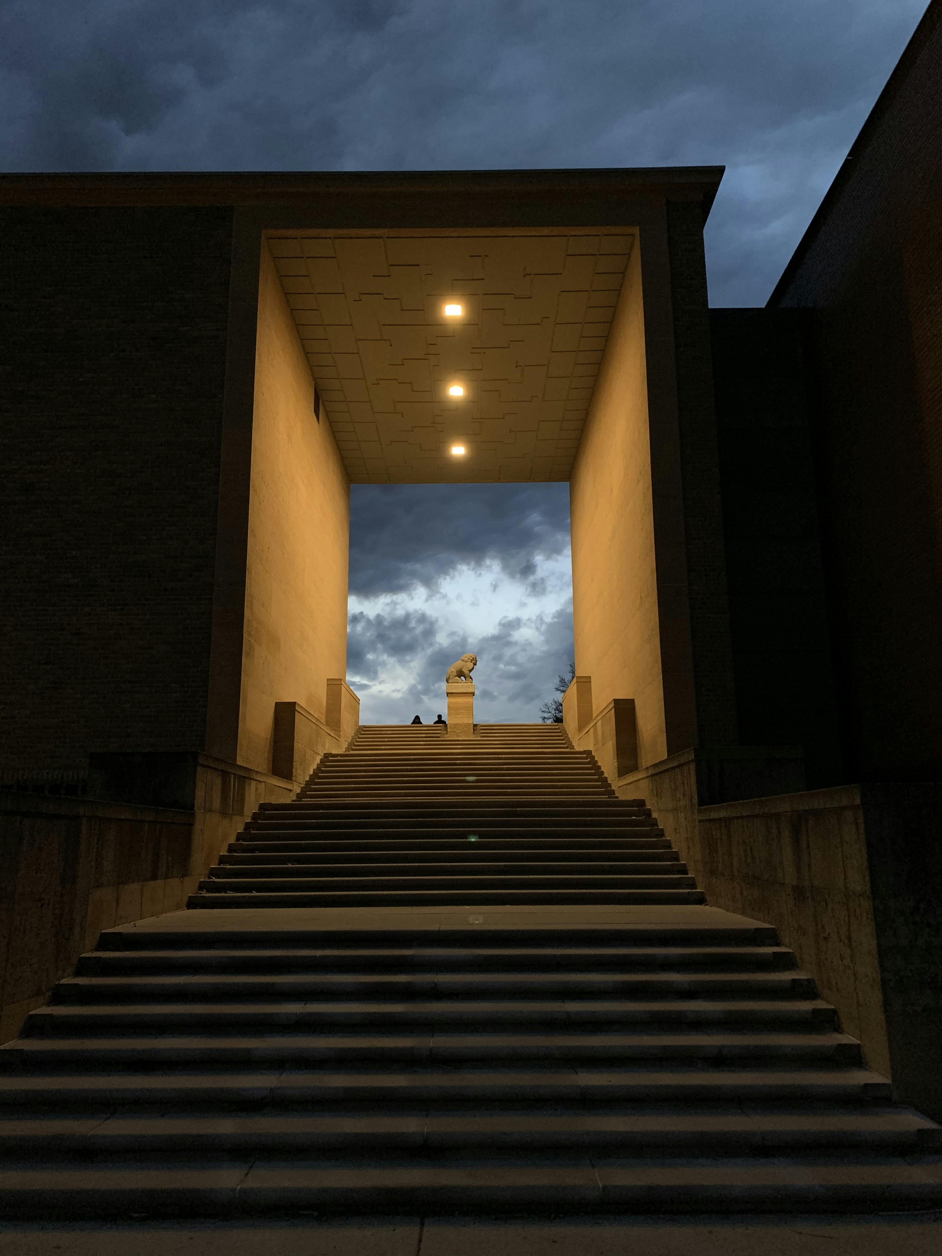 Illuminated staircase leading to a statue under a dramatic sky, framed by a modern architectural archway.