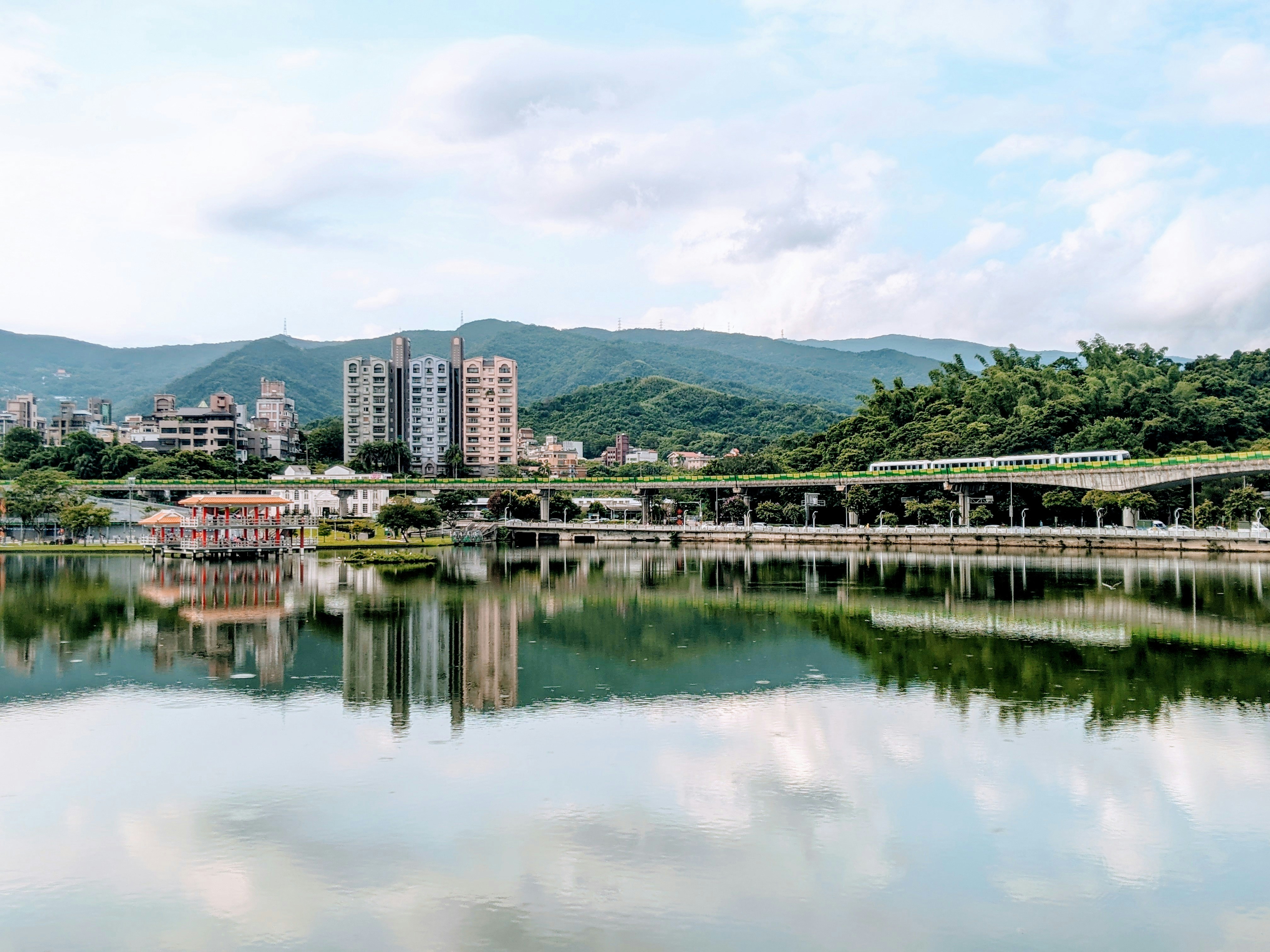 A tranquil lakeside view showcasing a train gliding above lush greenery, reflecting the surrounding buildings and mountains in the still water.