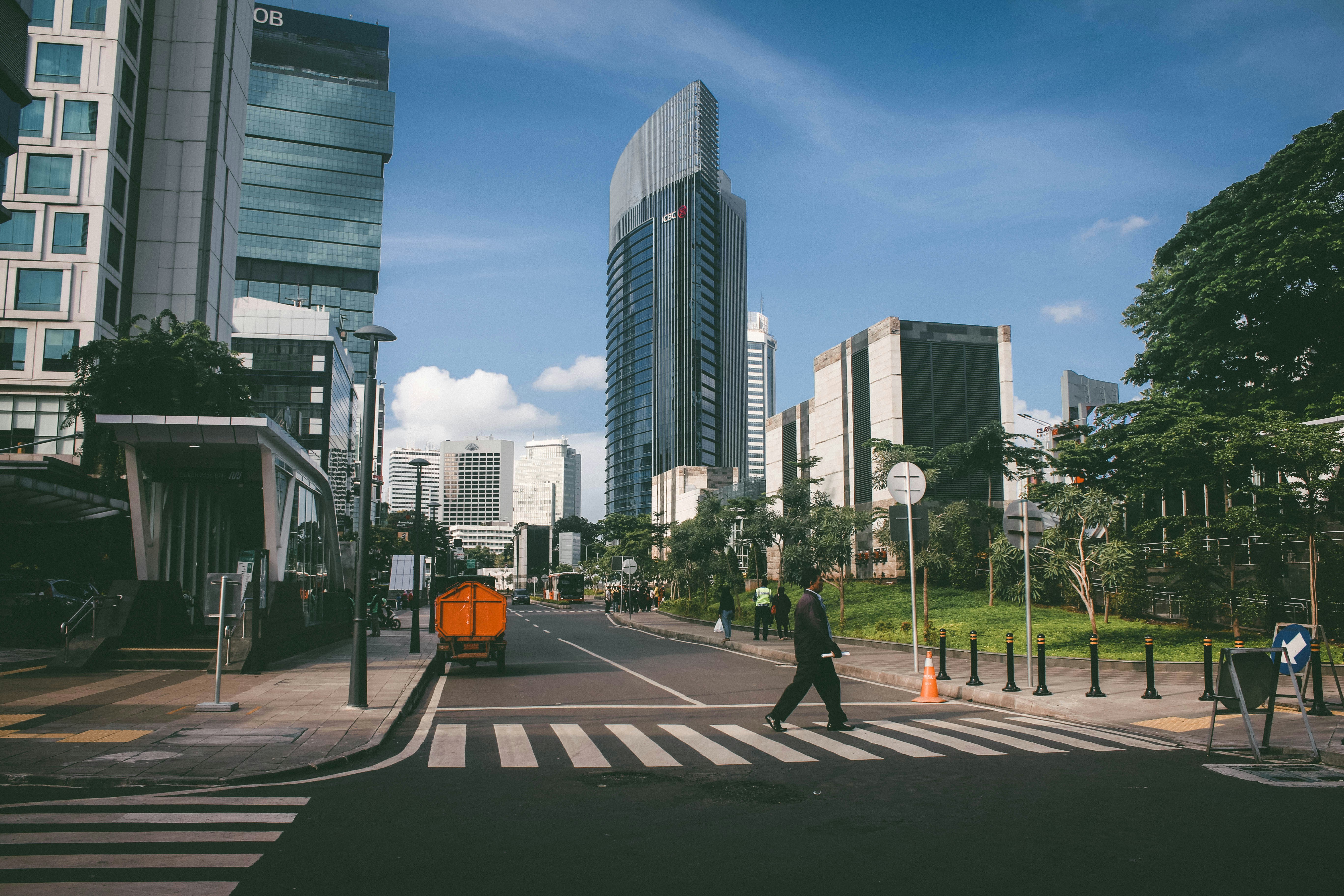 Businessman crossing a zebra-striped road in a bustling urban environment, flanked by modern skyscrapers and greenery.