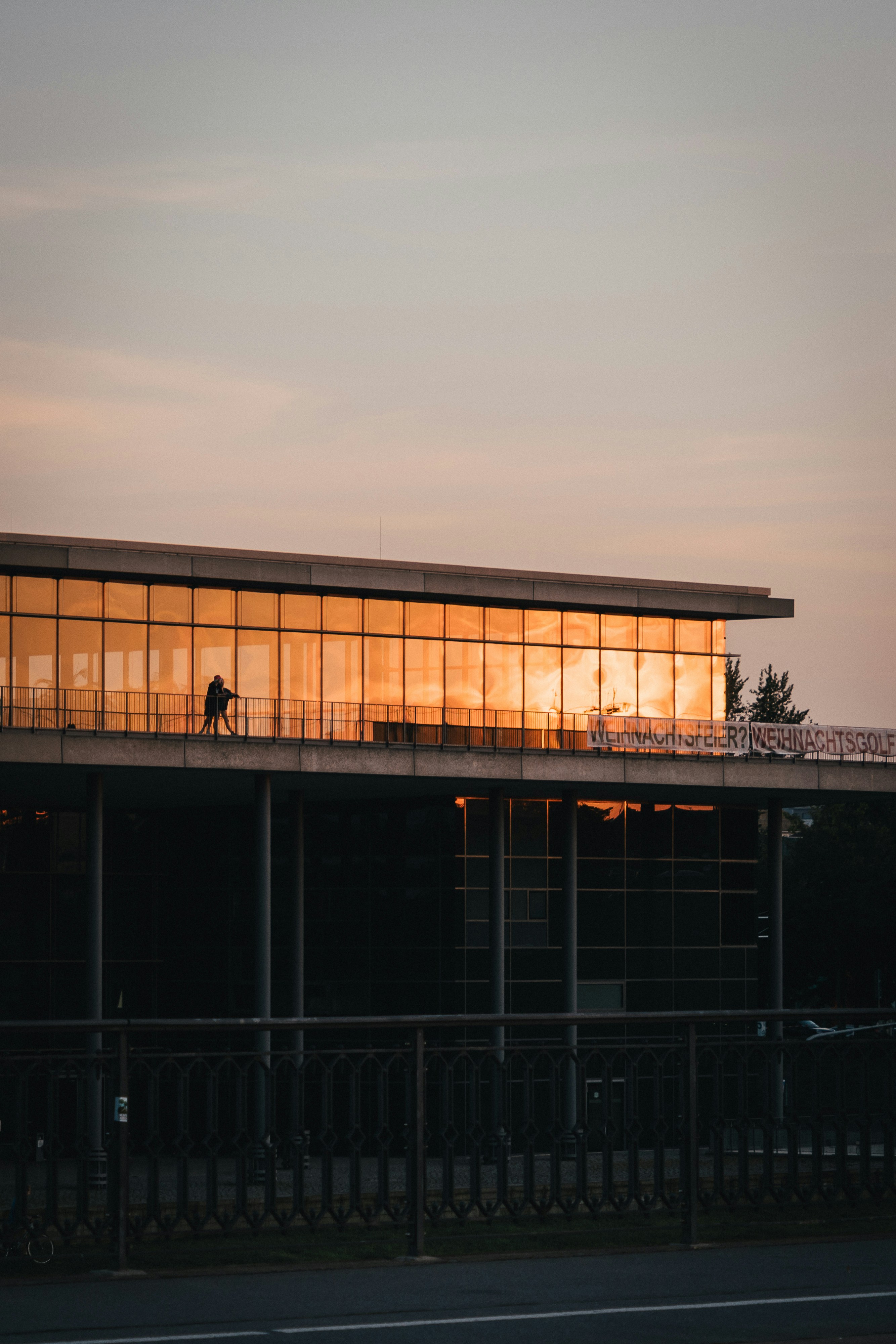 Sunset light bathes a glass-walled terrace of a modern building, with a lone figure standing on the balcony while a dark base and fence anchor the foreground.