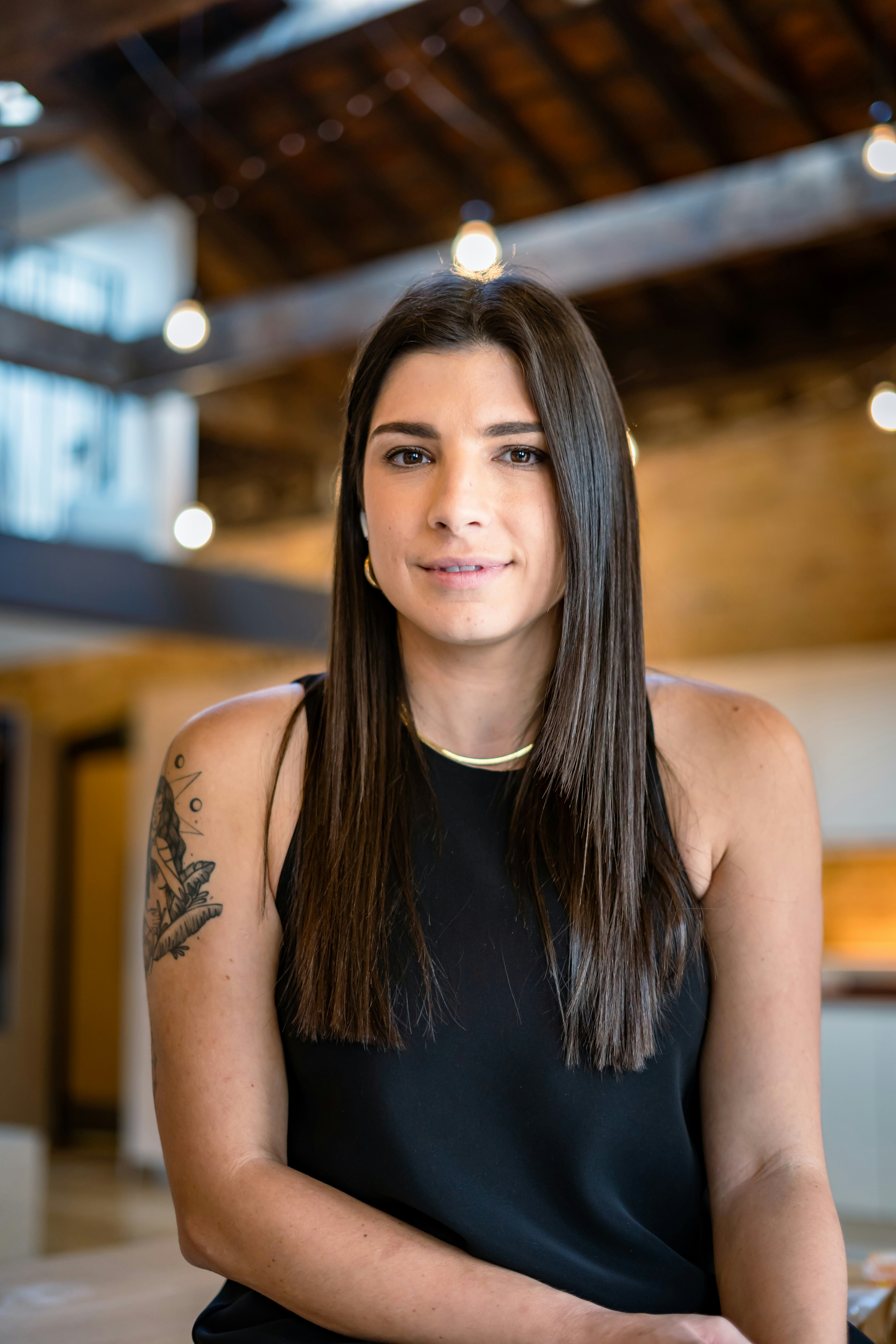 A poised individual with long hair and a tattoo, seated in a contemporary space featuring rustic wooden beams and soft lighting.