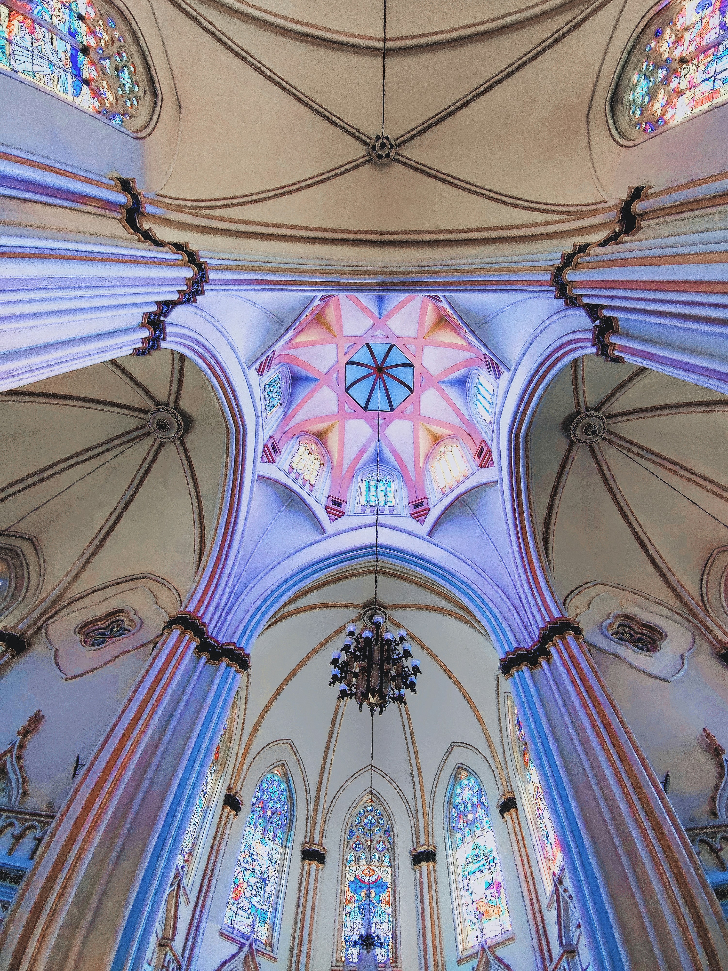 Intricate ceiling details of a grand church, showcasing ornate arches and colorful stained glass windows. A chandelier hangs elegantly at the center.
