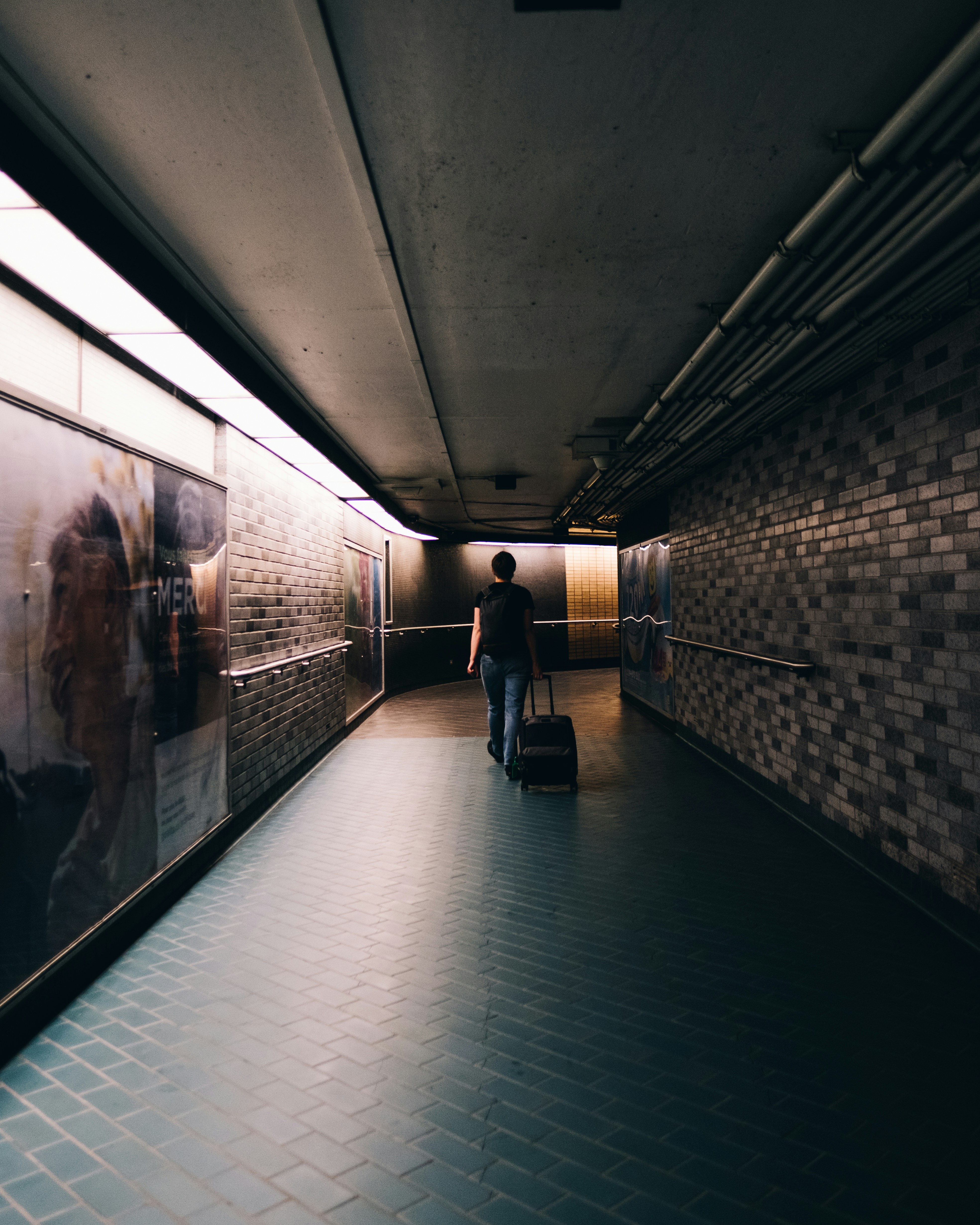 A traveler with a suitcase walks through a dimly lit subway corridor, flanked by colorful advertisements. The scene conveys a sense of solitude and anticipation.