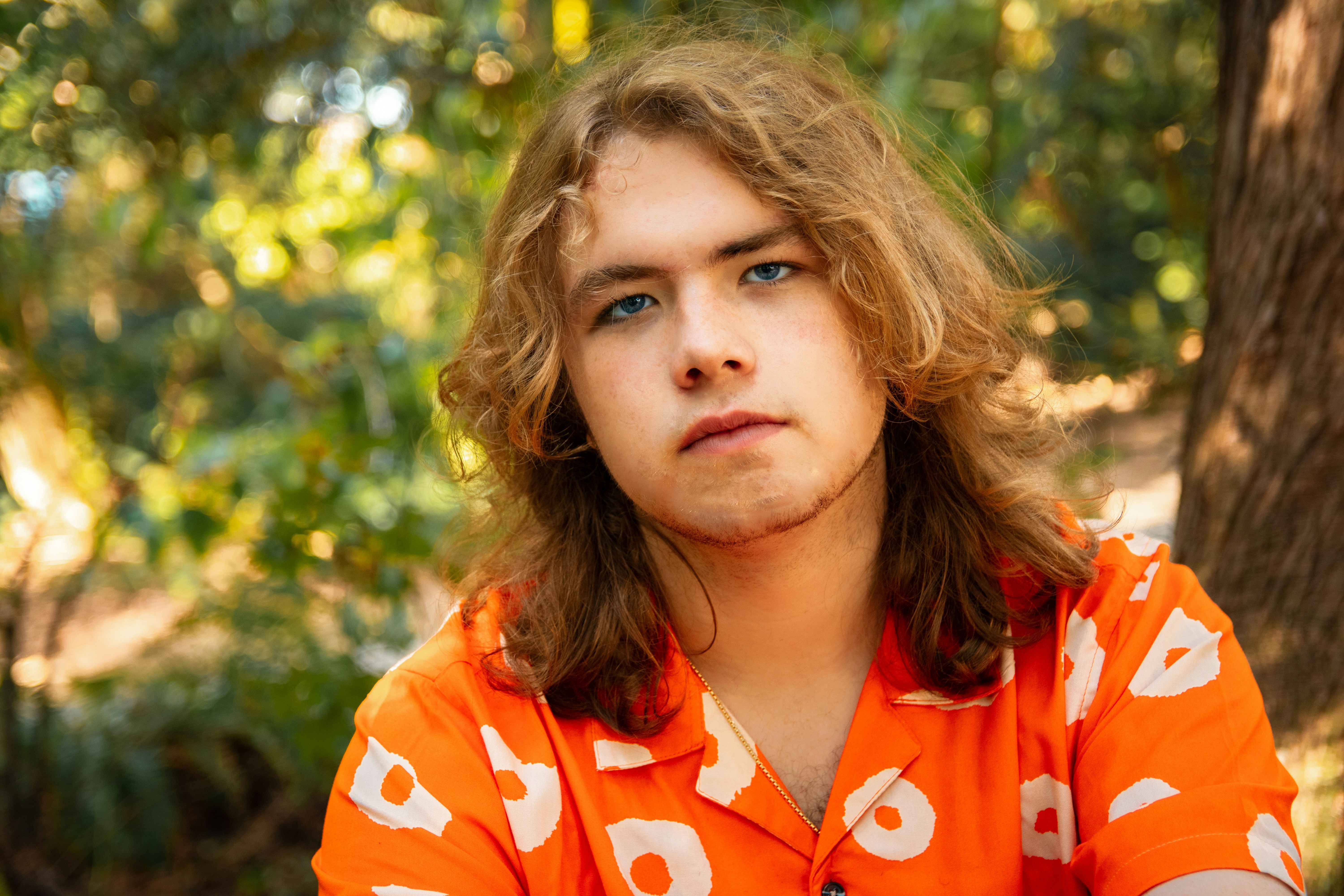 Young man with wavy hair wearing a bright orange shirt sits thoughtfully amidst lush greenery. The warm colors create an inviting atmosphere.