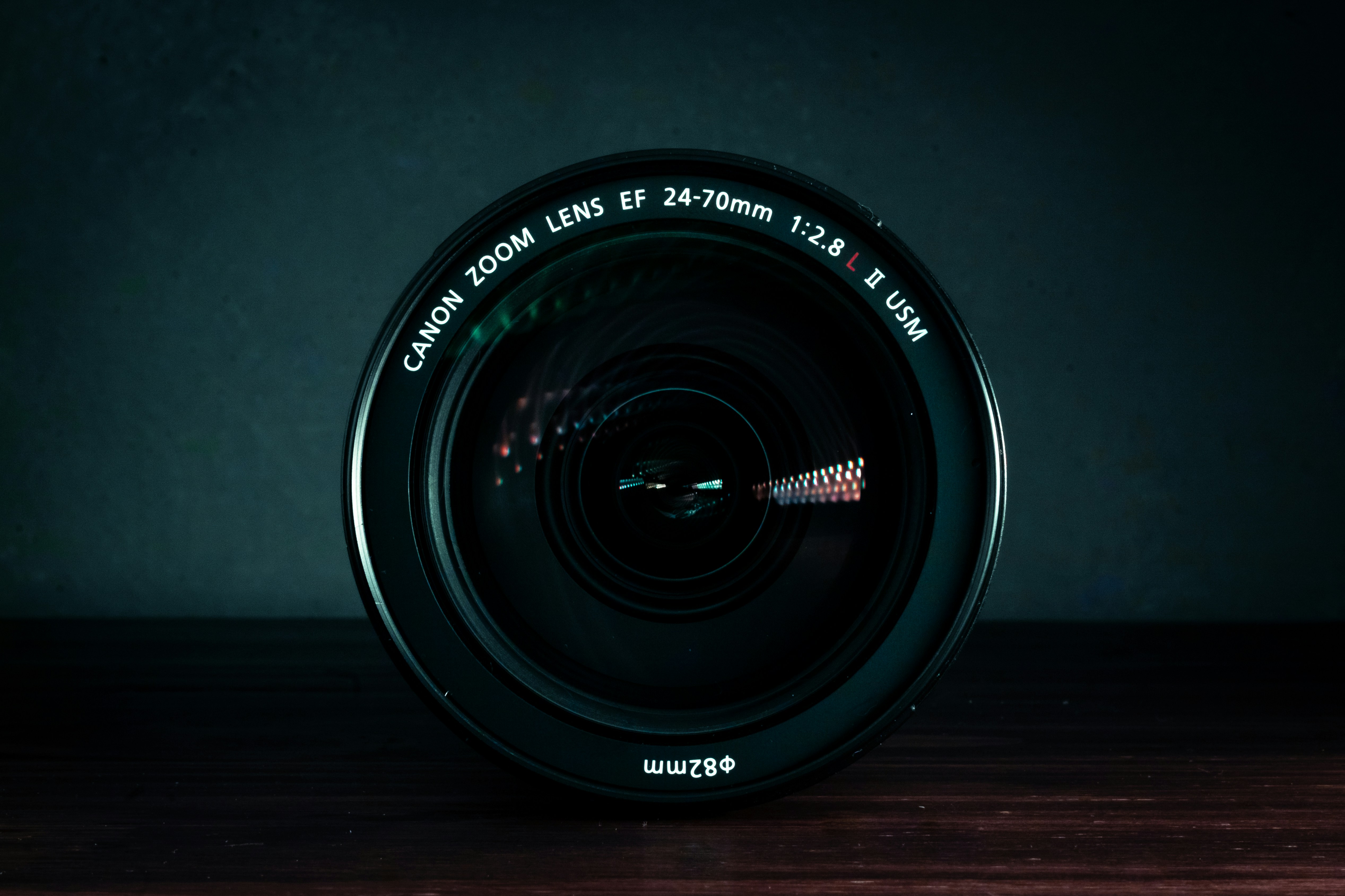 Canon Zoom Lens EF 24-70mm f/2.8 II USM resting on a wooden surface with a dark background, showcasing its sleek design and precision optics.