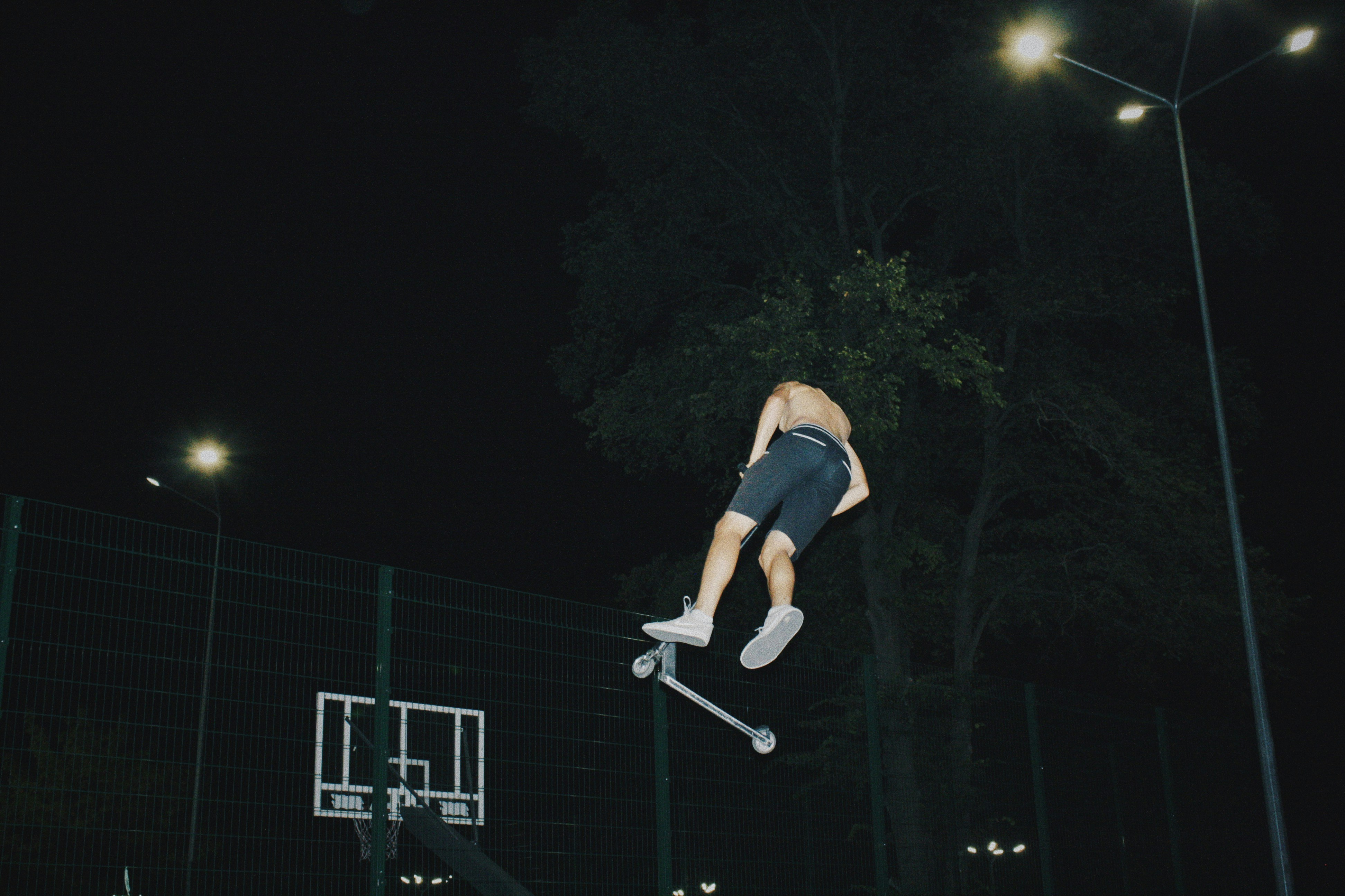 Scooter rider performing an aerial trick against a backdrop of a dimly lit basketball court at night.