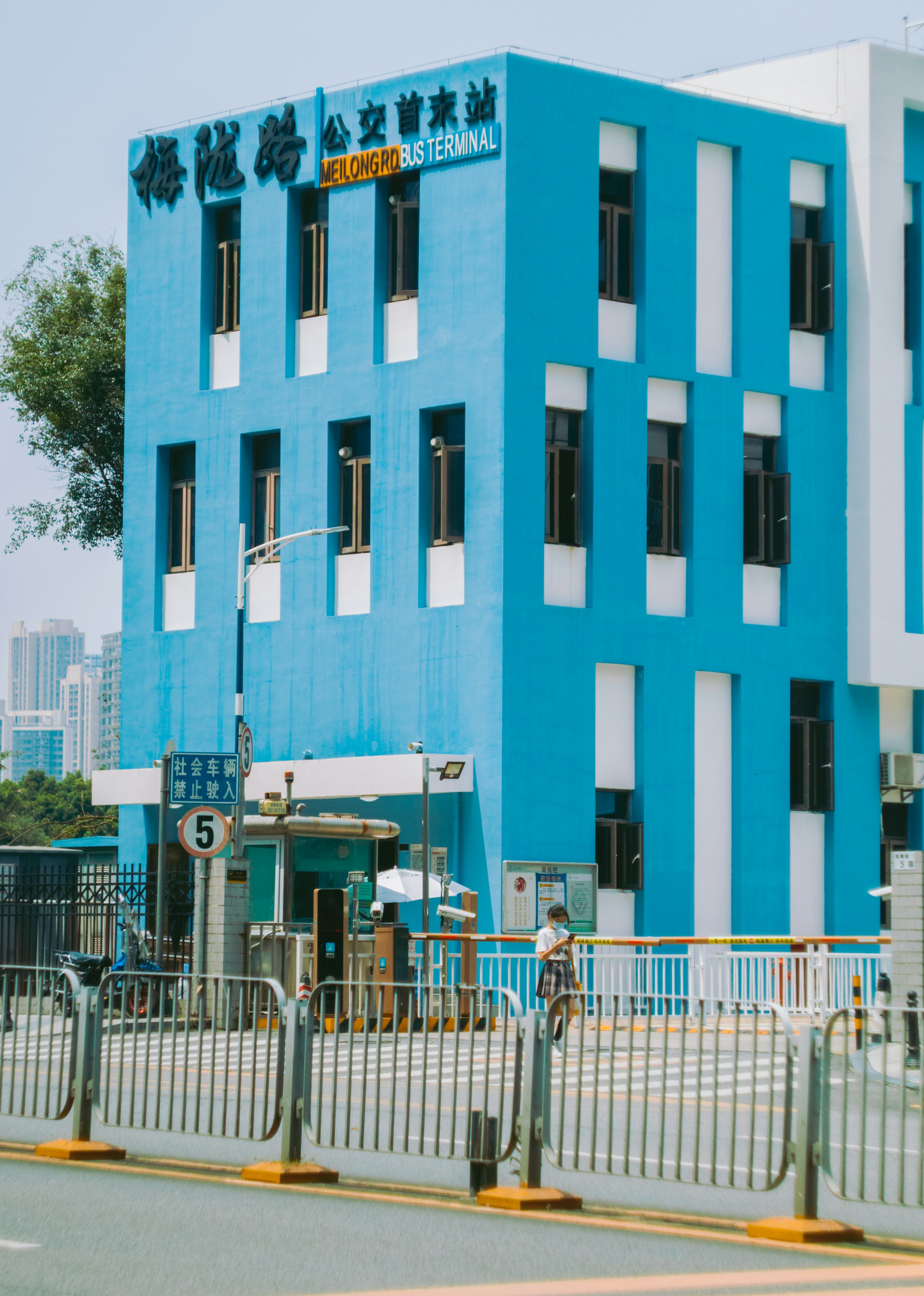 Vibrant blue bus terminal with modern architecture, featuring large windows and clear signage. A pedestrian navigates the area, adding life to the urban scene.