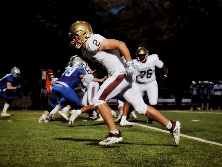 High-school football player wearing #2 sprinting off the line amid linemen under stadium lights.