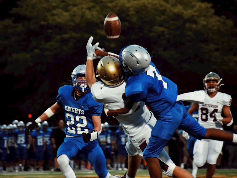 High-school football players collide midair as a loose ball arcs above them during a night game.