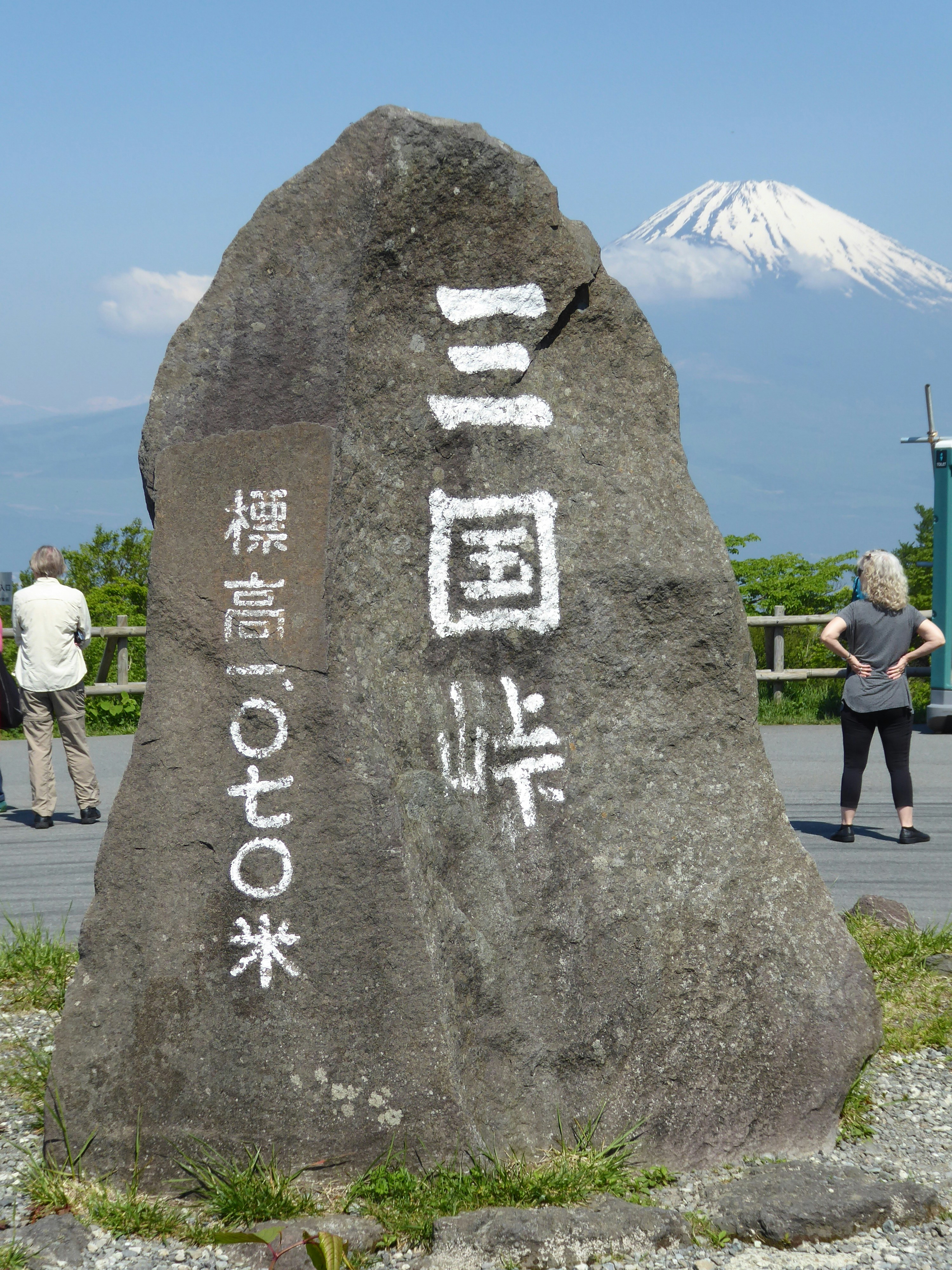 A large rock formation with inscriptions stands prominently, framed by a distant view of snow-capped Mount Fuji. Visitors admire the scenic landscape.