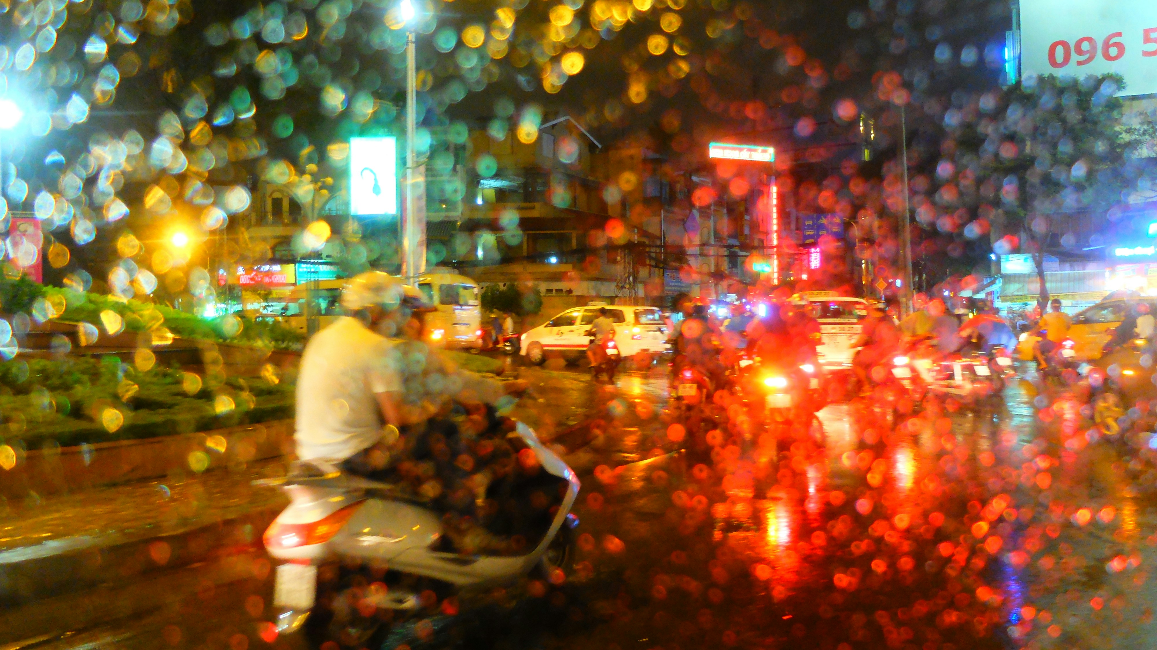 Night street scene with a rider on a scooter amid neon headlights and rain-kissed reflections.