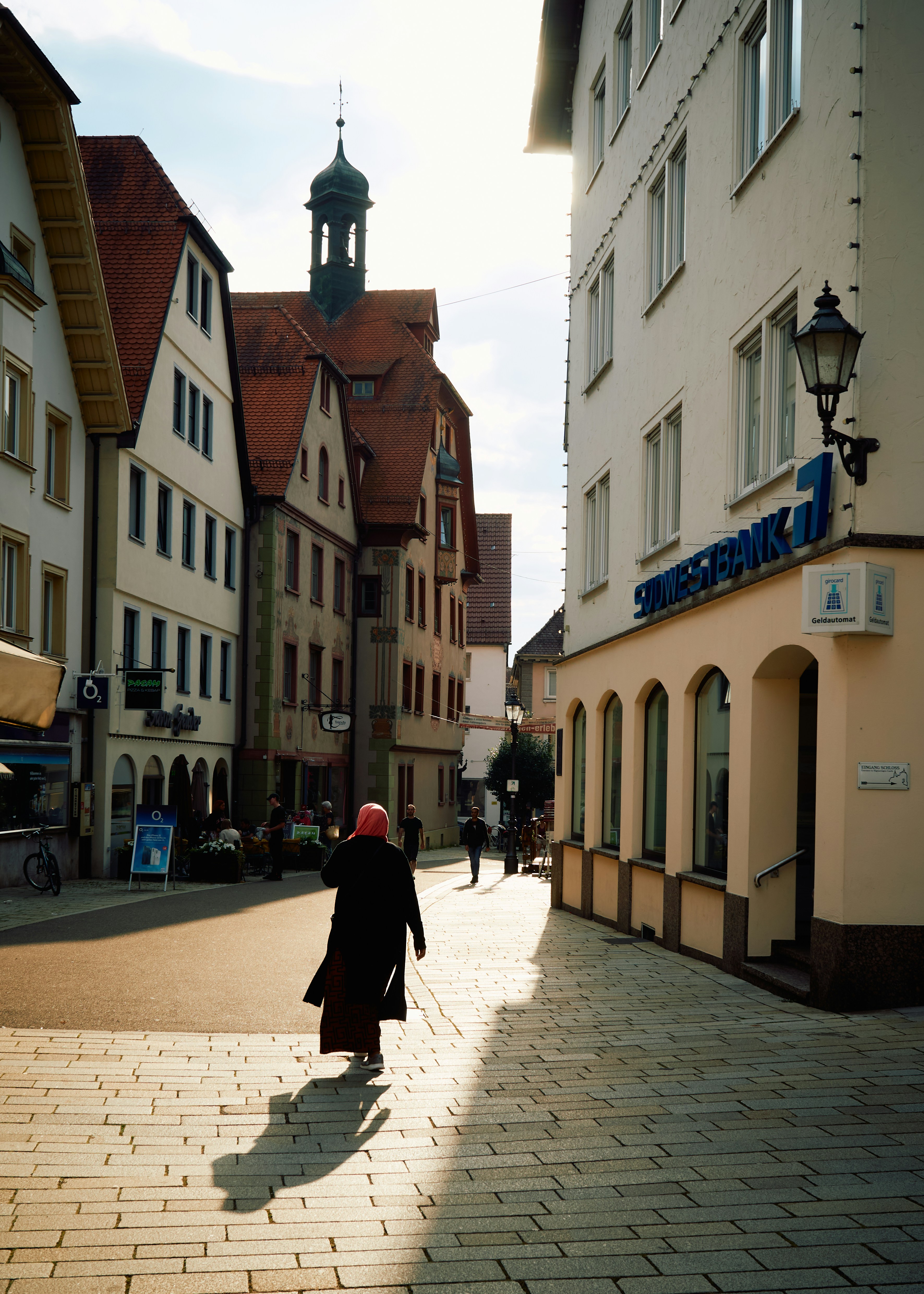 Sunlit Street Corridor: A Pedestrian Walks Toward a Green-Domed Clock Tower