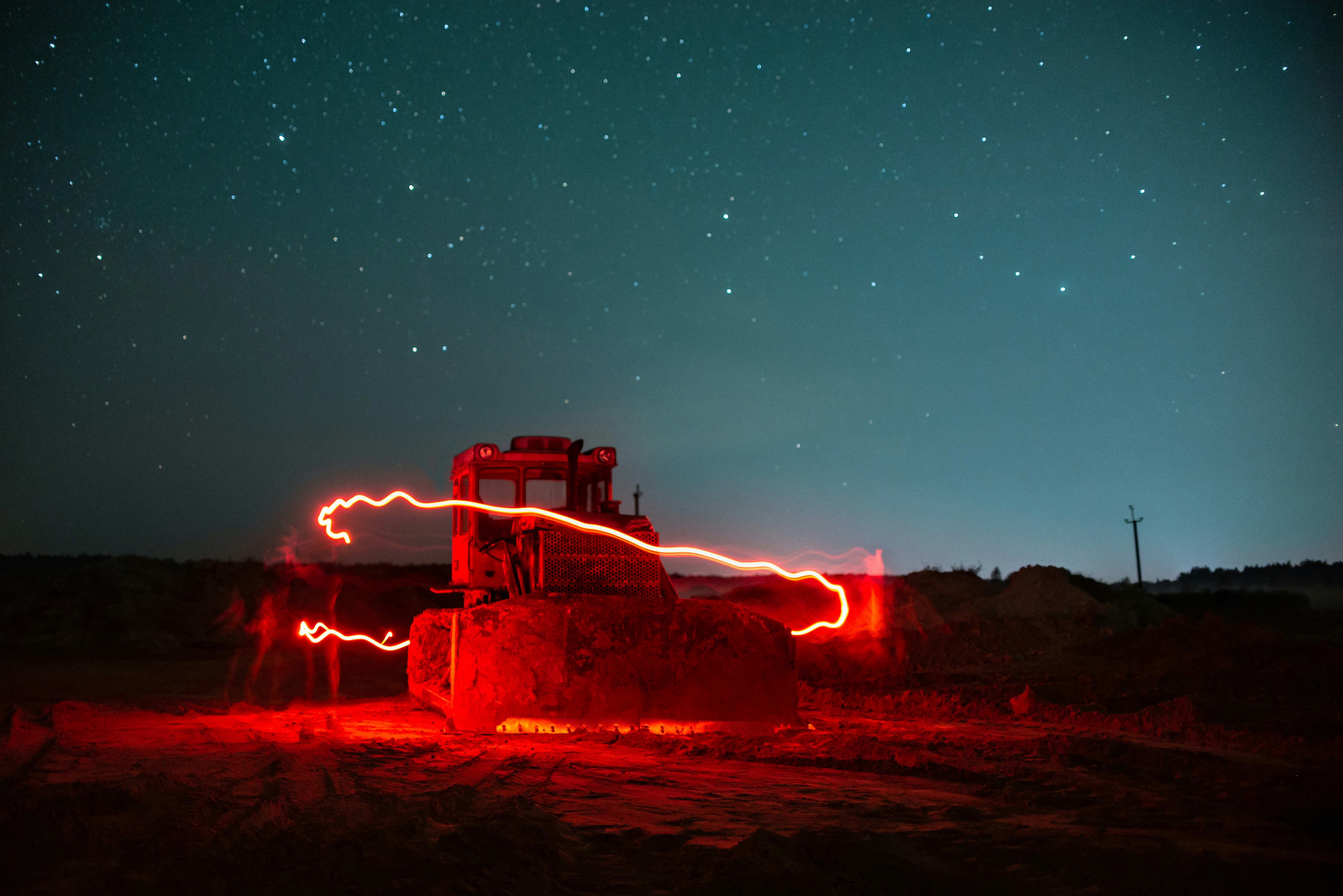 A red-lit bulldozer sits in a dark landscape, illuminated by trails of light against a starry sky. The scene captures the juxtaposition of industrial presence and celestial beauty.