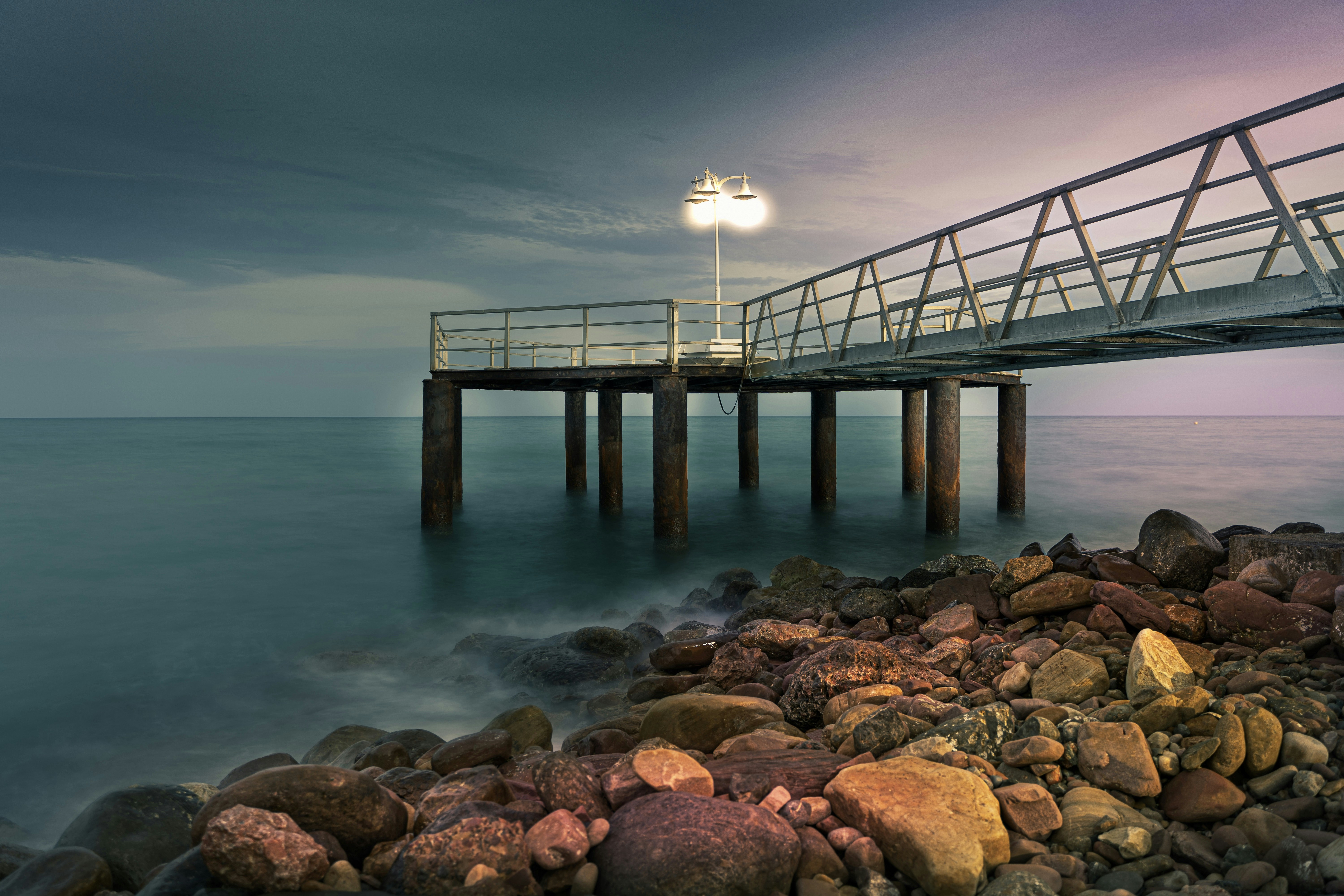 A serene pier extends over calm waters, illuminated by a glowing lamp against a moody sky. Rocky shoreline adds texture to the tranquil scene.