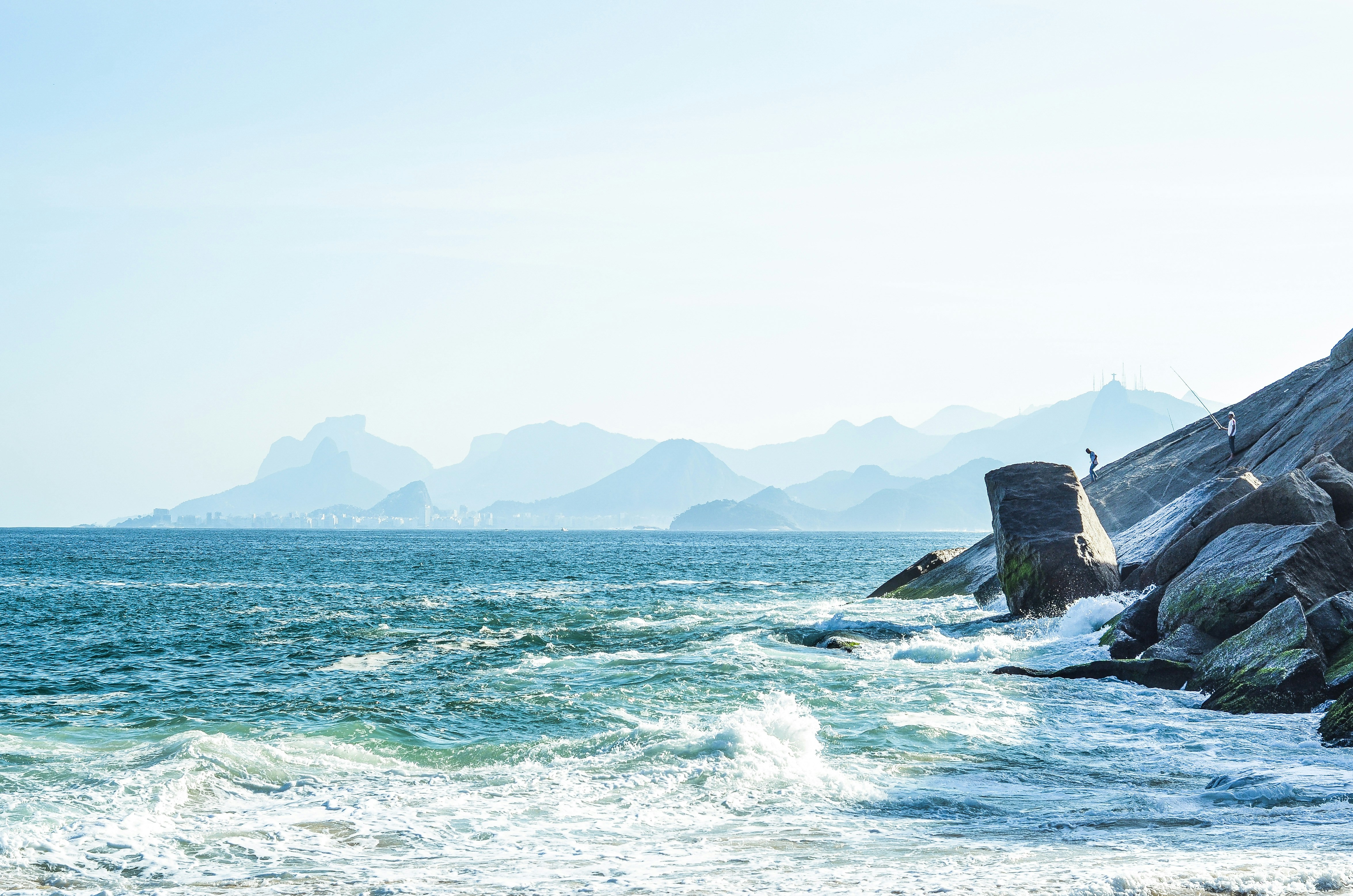 Waves crashing against a rocky shoreline with distant mountains under a clear blue sky.