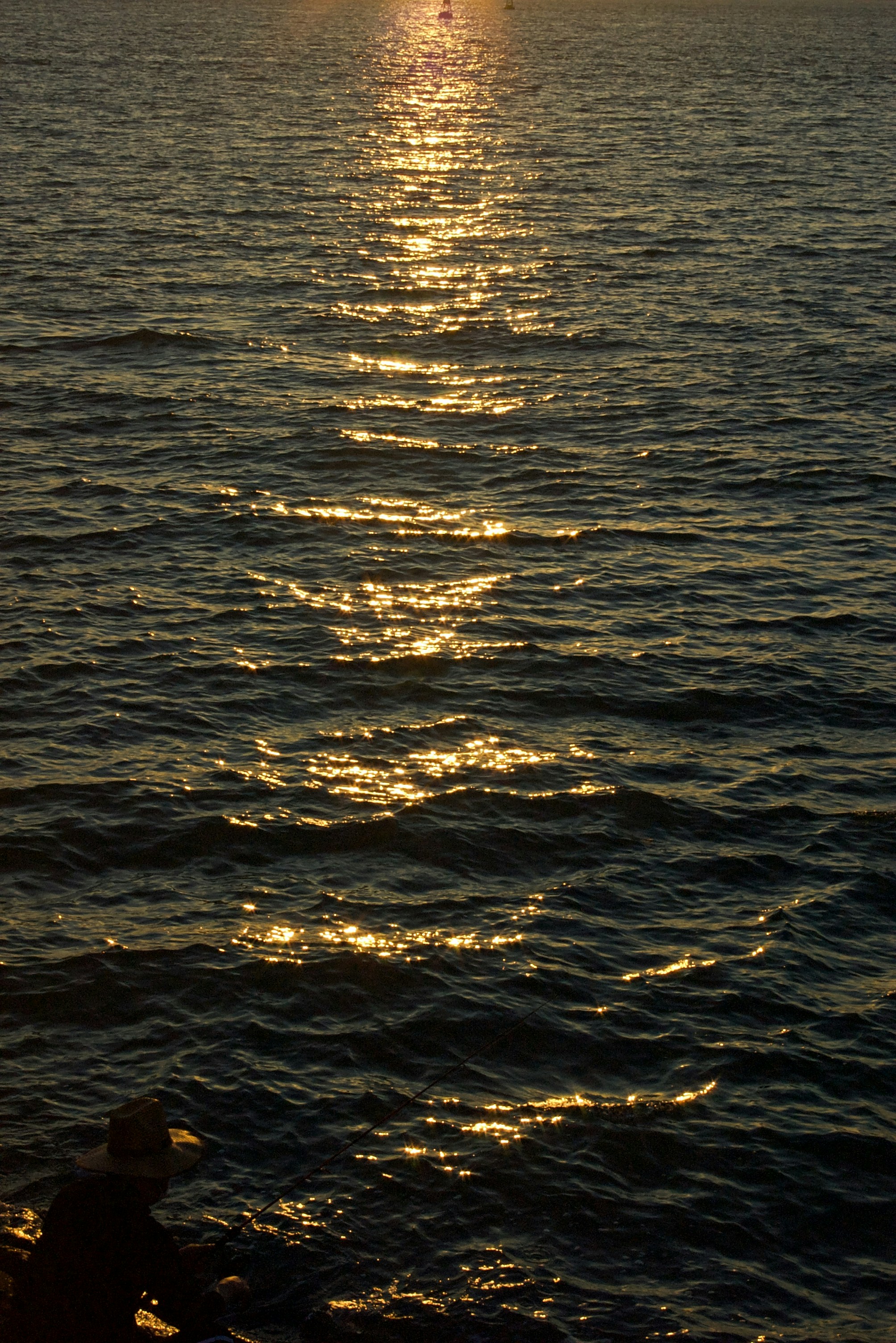 Sunlight shimmering on the surface of a calm sea at dusk, with a figure in a hat engaged in fishing. 
