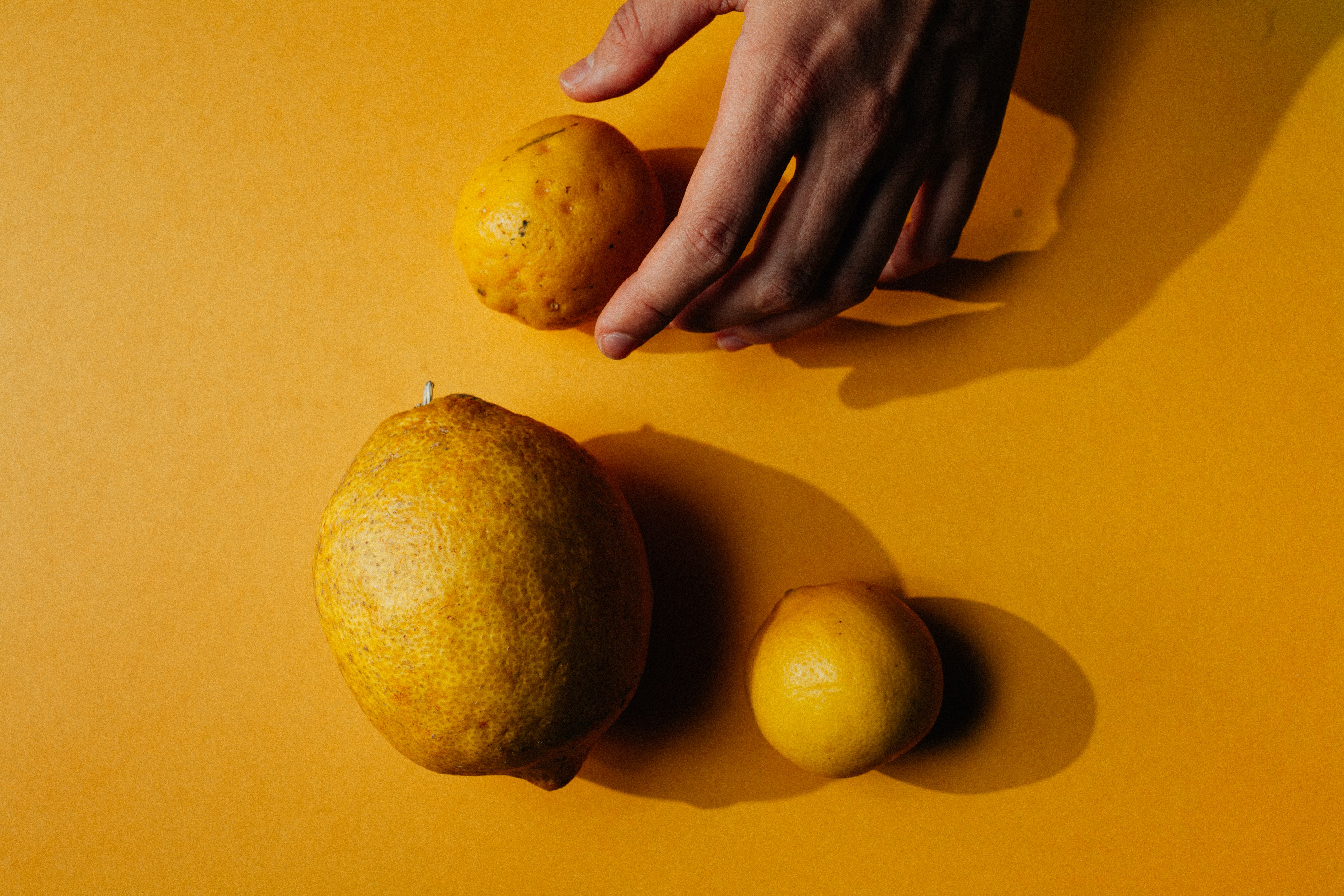 A hand reaches for a lemon among a trio of citrus fruits against a vibrant orange background, emphasizing texture and form.