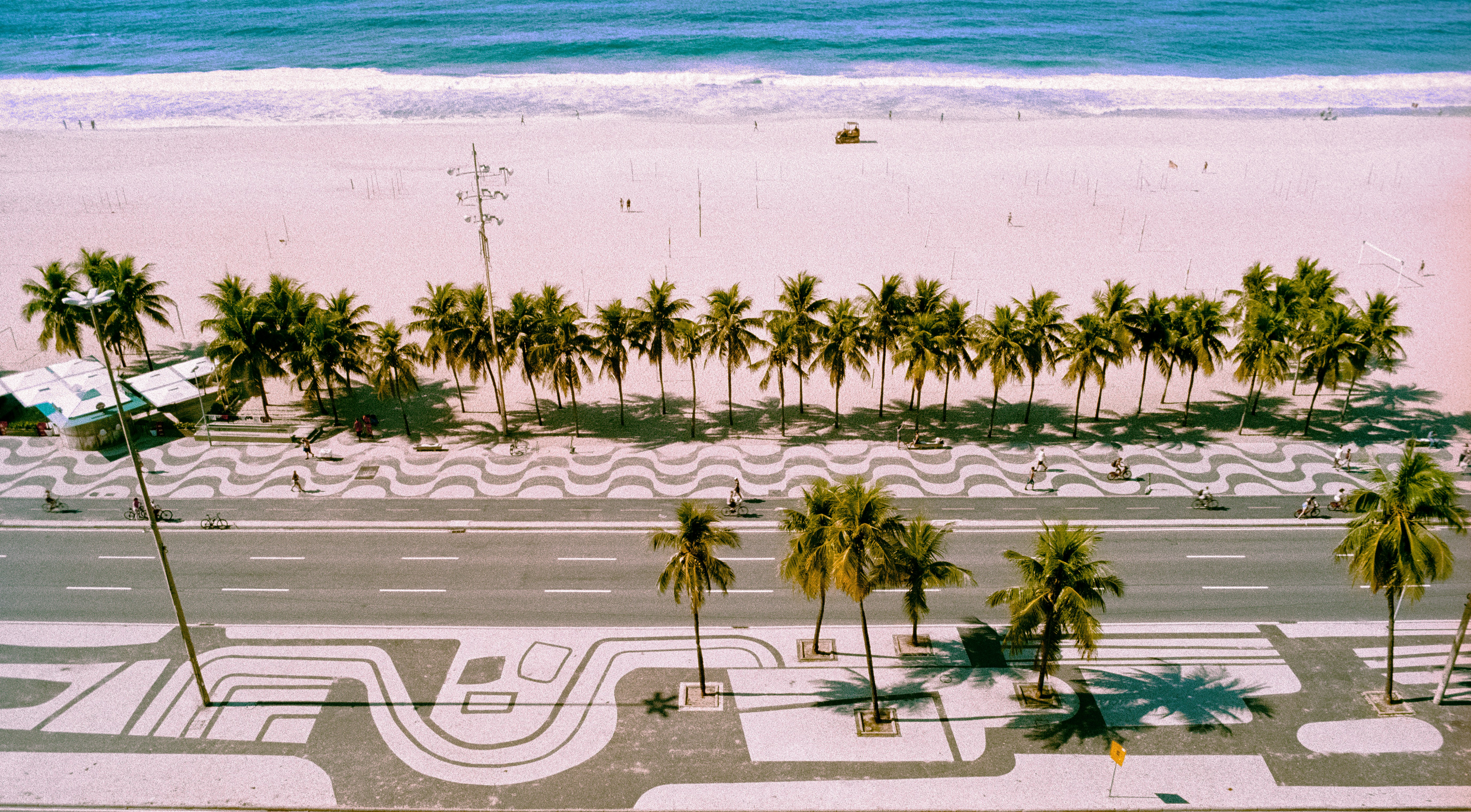 Aerial photograph of a seaside promenade where palm trees line a patterned, geometric plaza beside a sandy beach and turquoise sea.