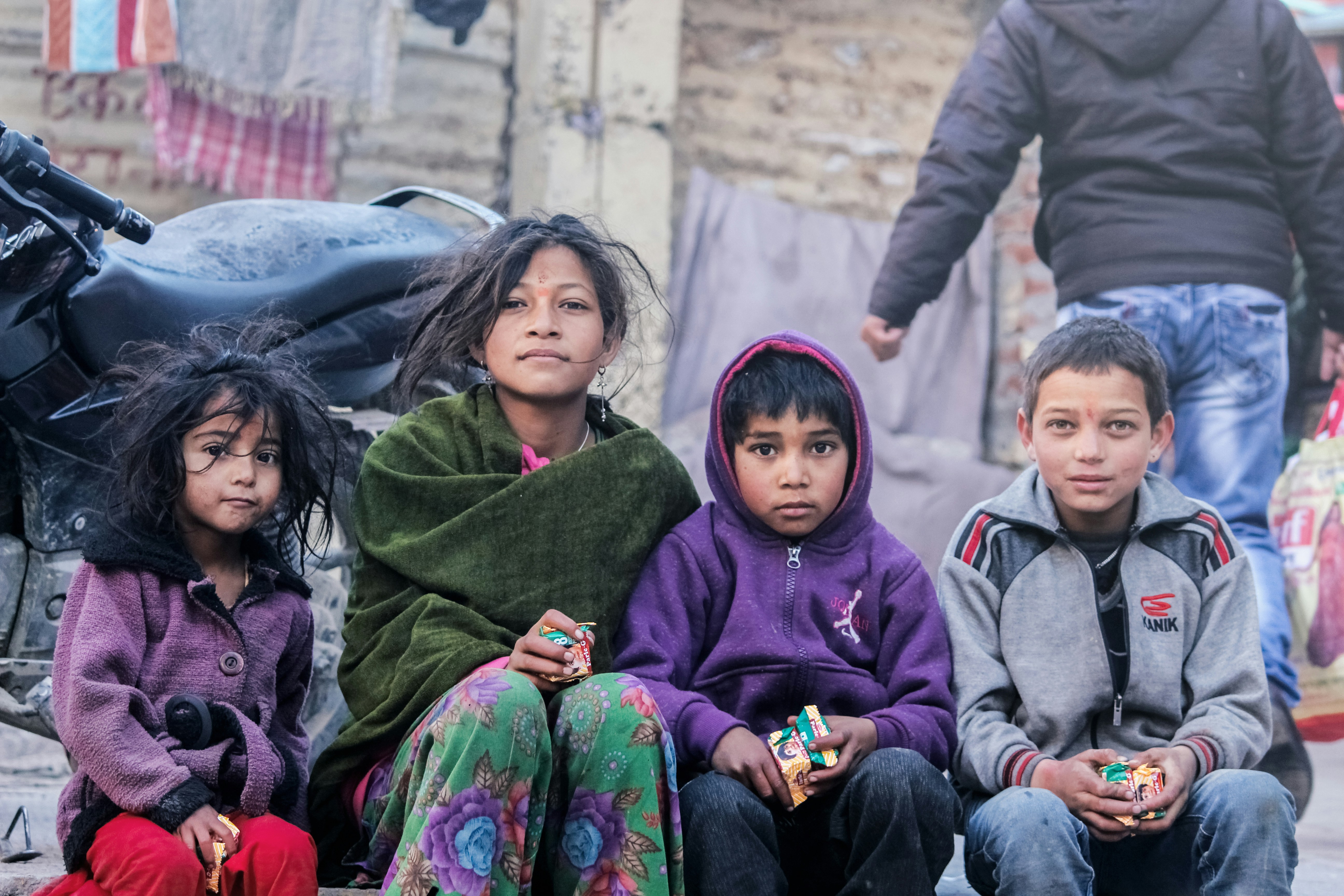 Four children bundled up in warm clothing sit together, holding snacks in an urban setting.