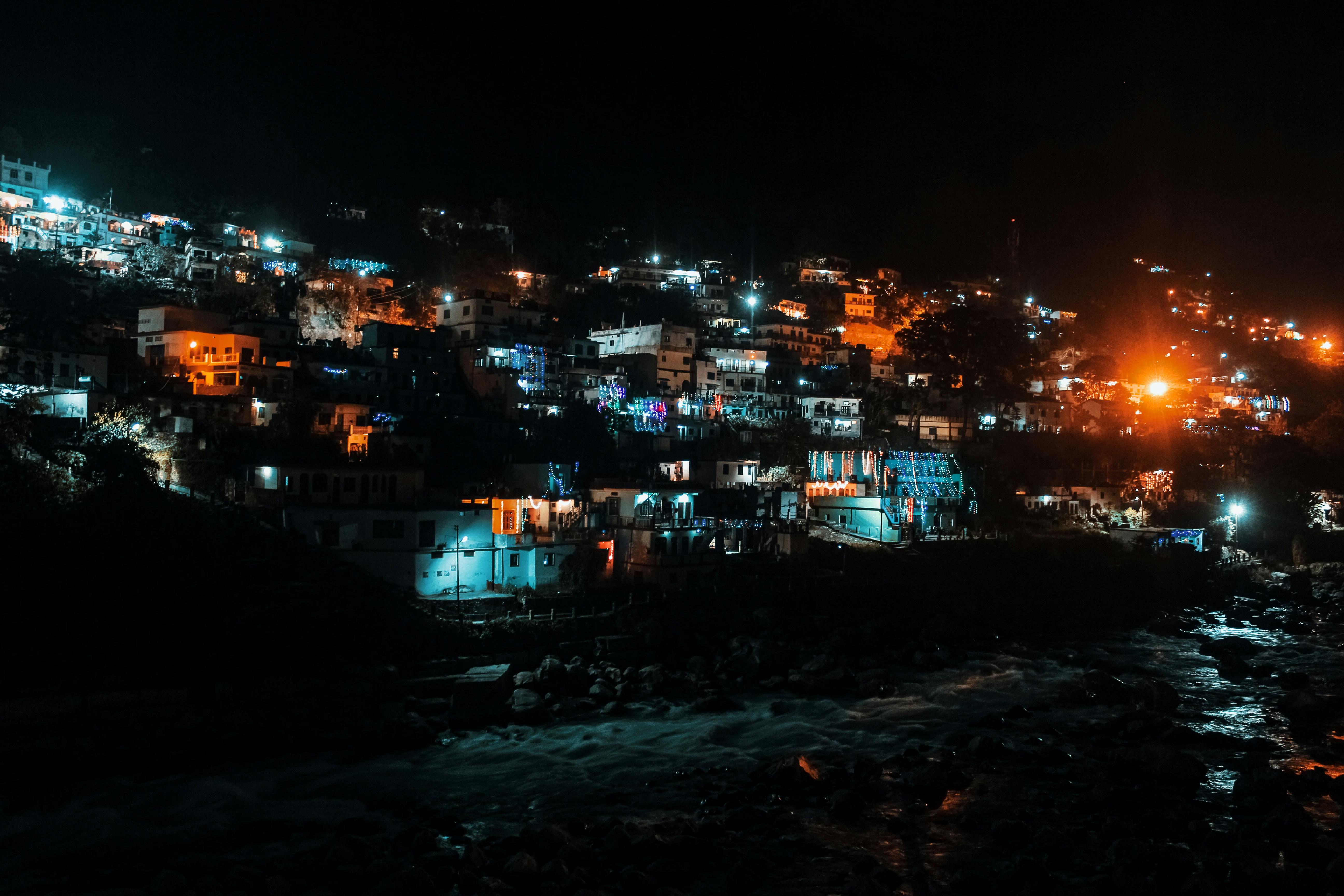 Vibrant hillside village illuminated by colorful lights at night, reflecting the lively atmosphere and urban charm. The flowing river adds a serene contrast to the bustling scene.