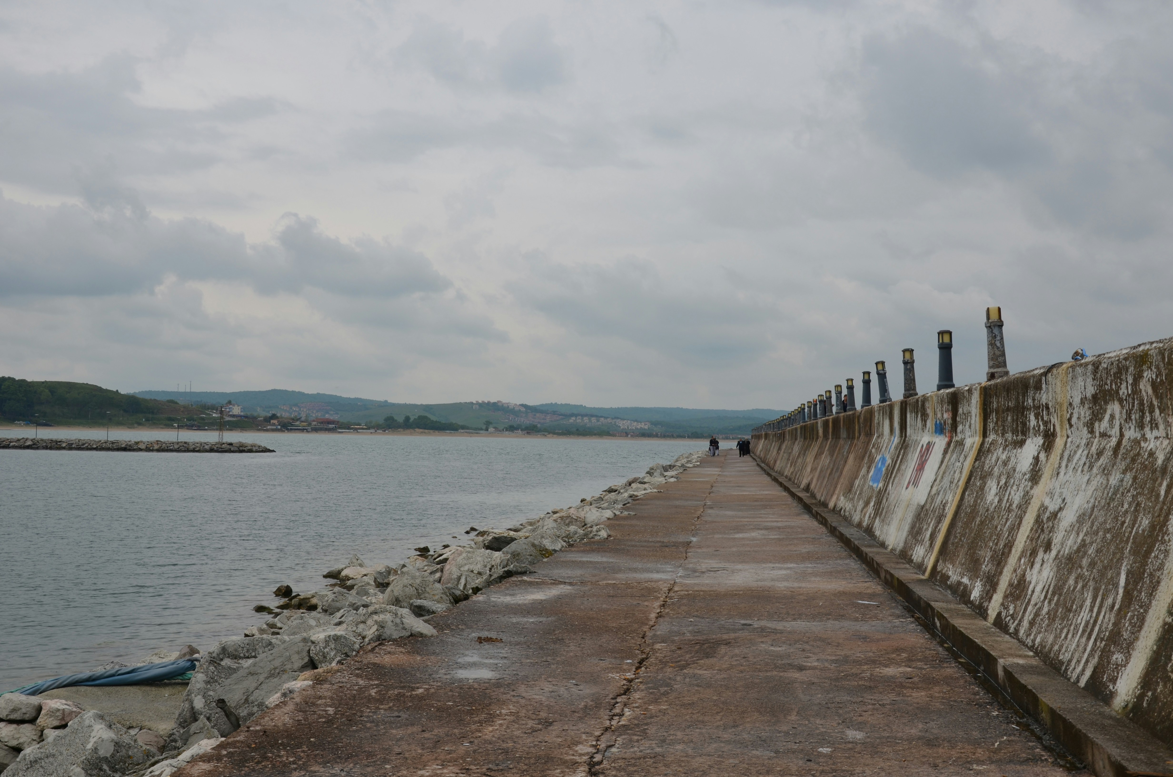 A long, weathered jetty extends into a calm sea under a cloudy sky, with distant hills visible on the horizon.