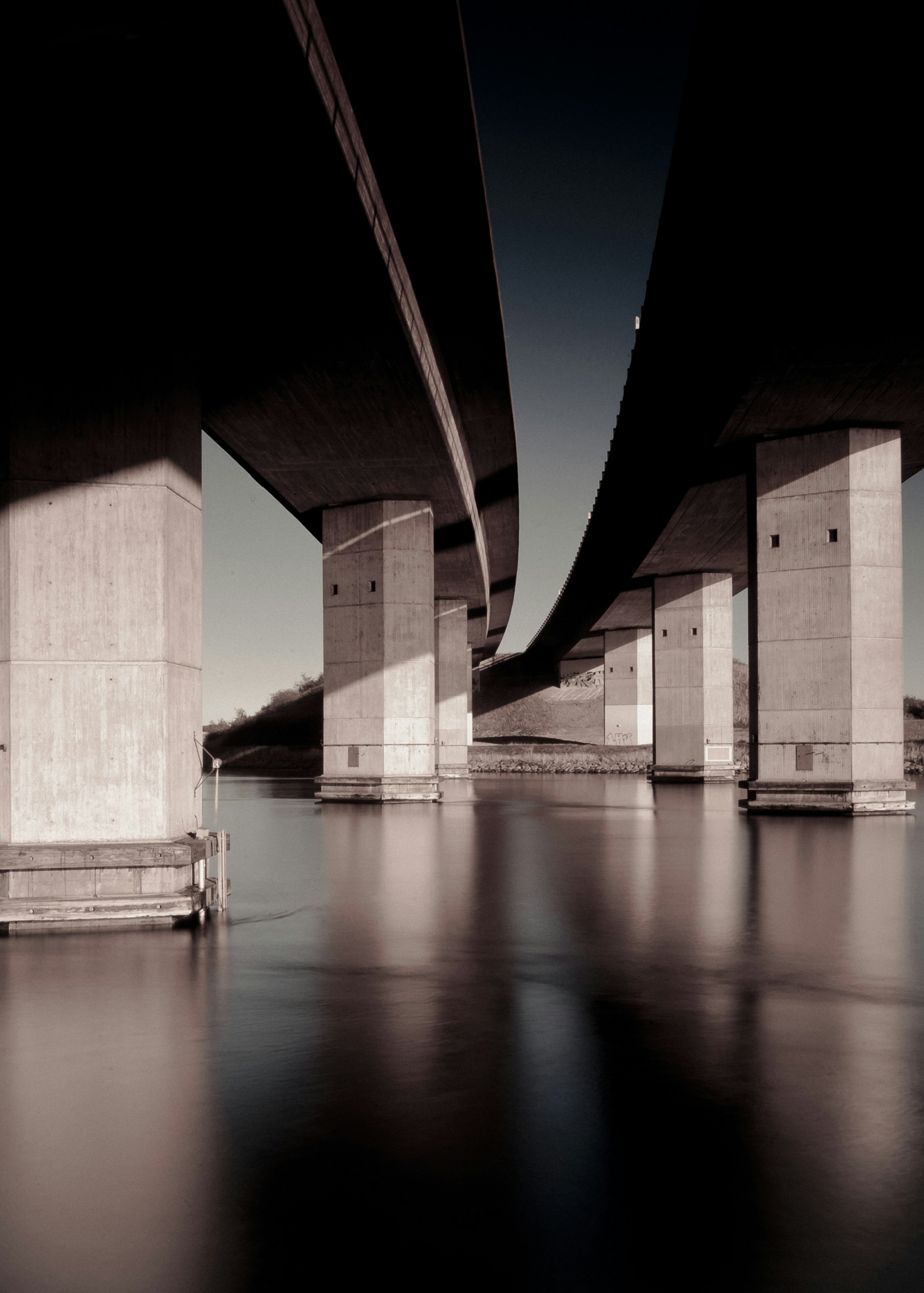 Concrete pillars of a bridge rise above calm waters, creating a striking interplay of light and shadow.