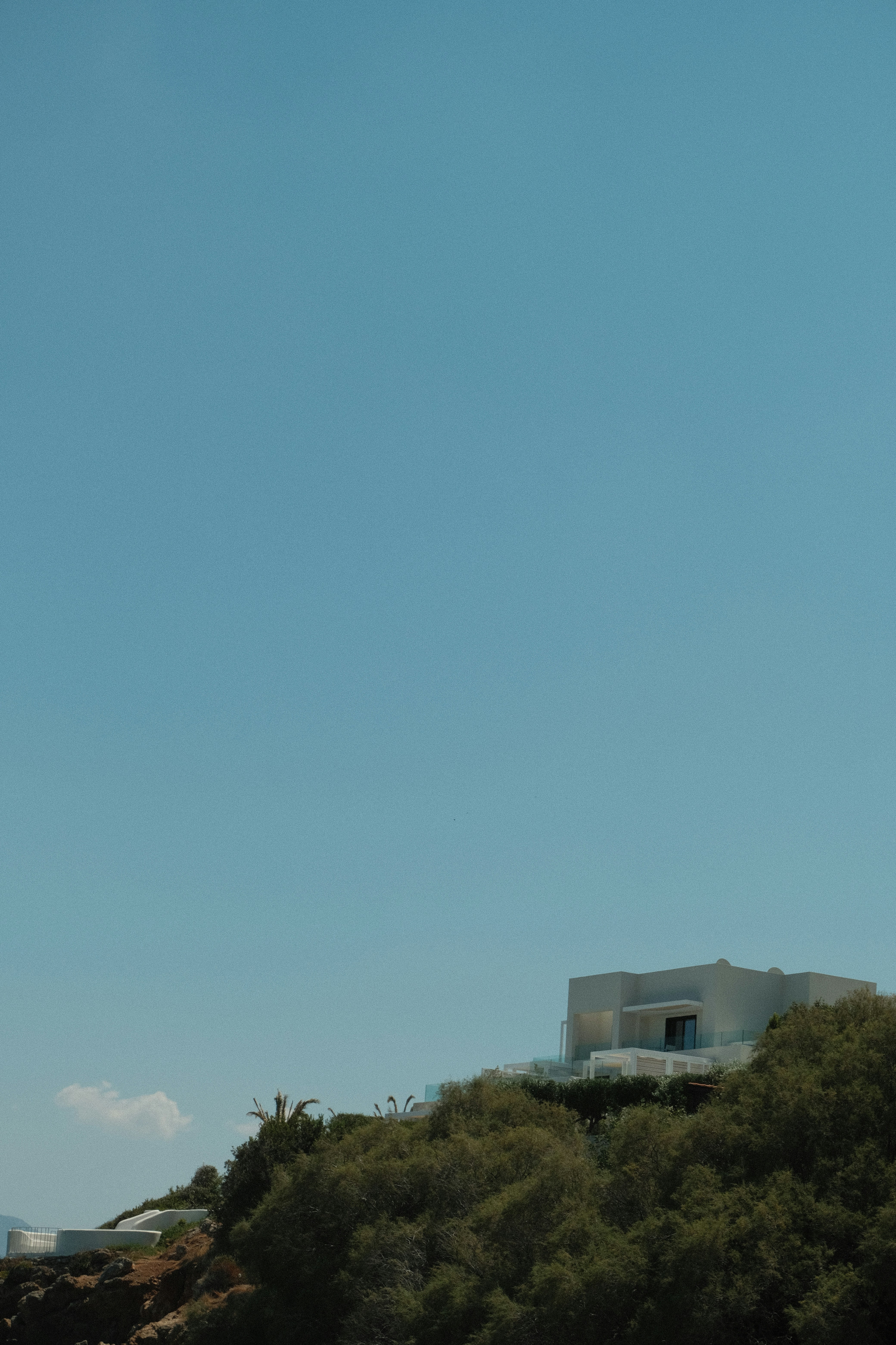 Modern white structure perched on a hillside, surrounded by lush greenery under a clear blue sky.