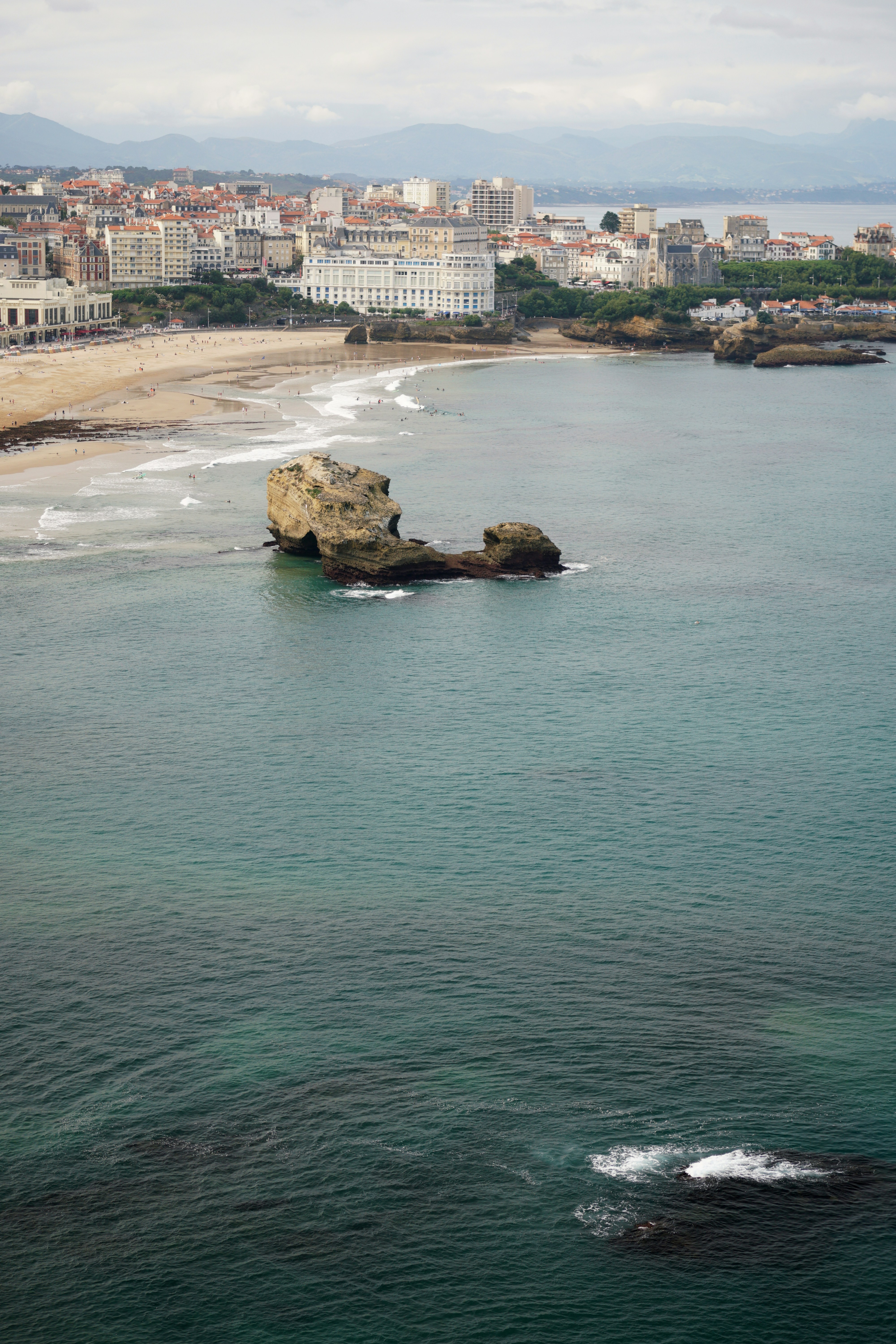 Rock formations jutting out from the turquoise waters, with a vibrant beach and townscape in the background. Coastal scene captures the blend of nature and urban life.