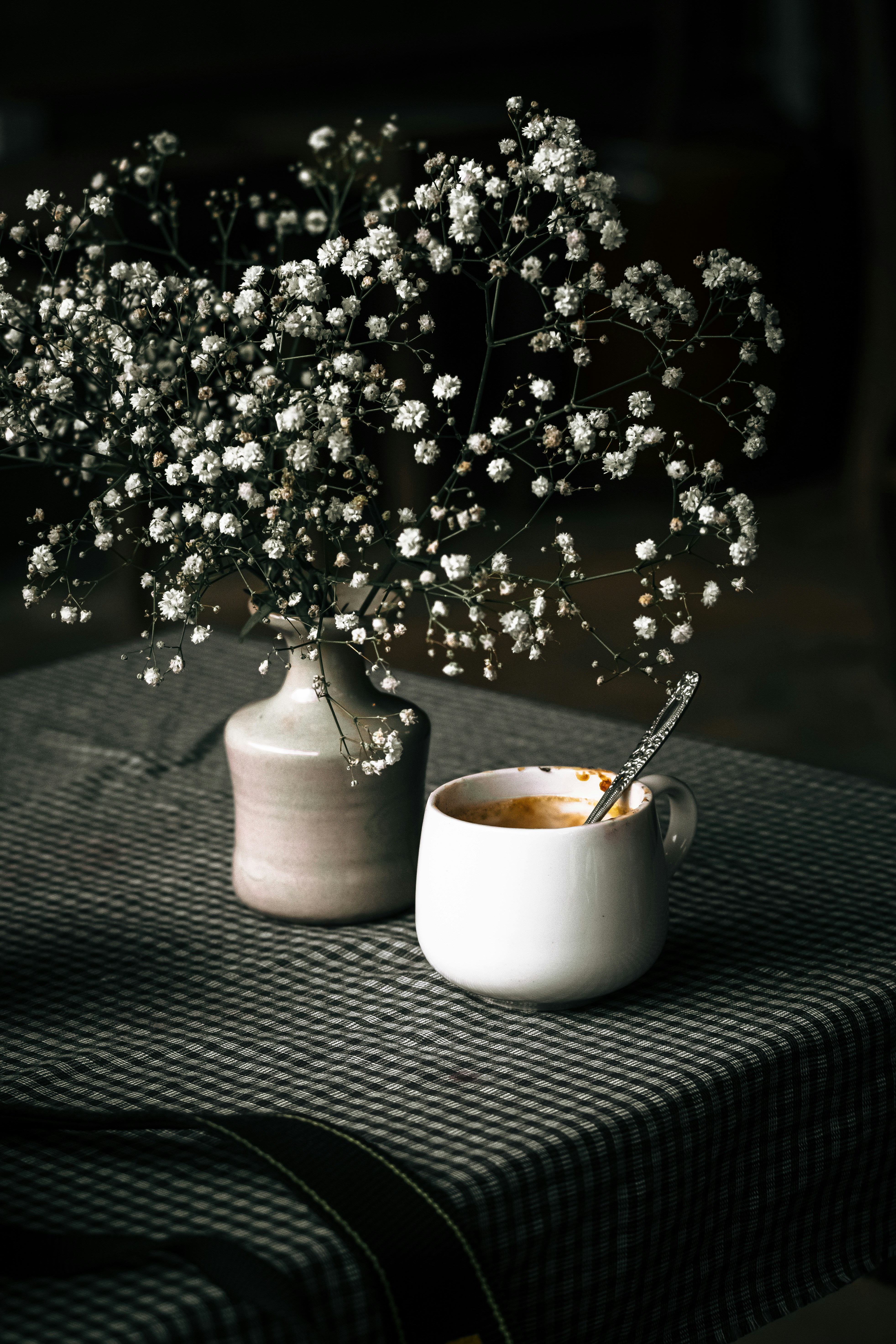 Still-life photograph of a delicate baby's breath bouquet in a ceramic vase beside a white cup of coffee on a dark checkered tablecloth.