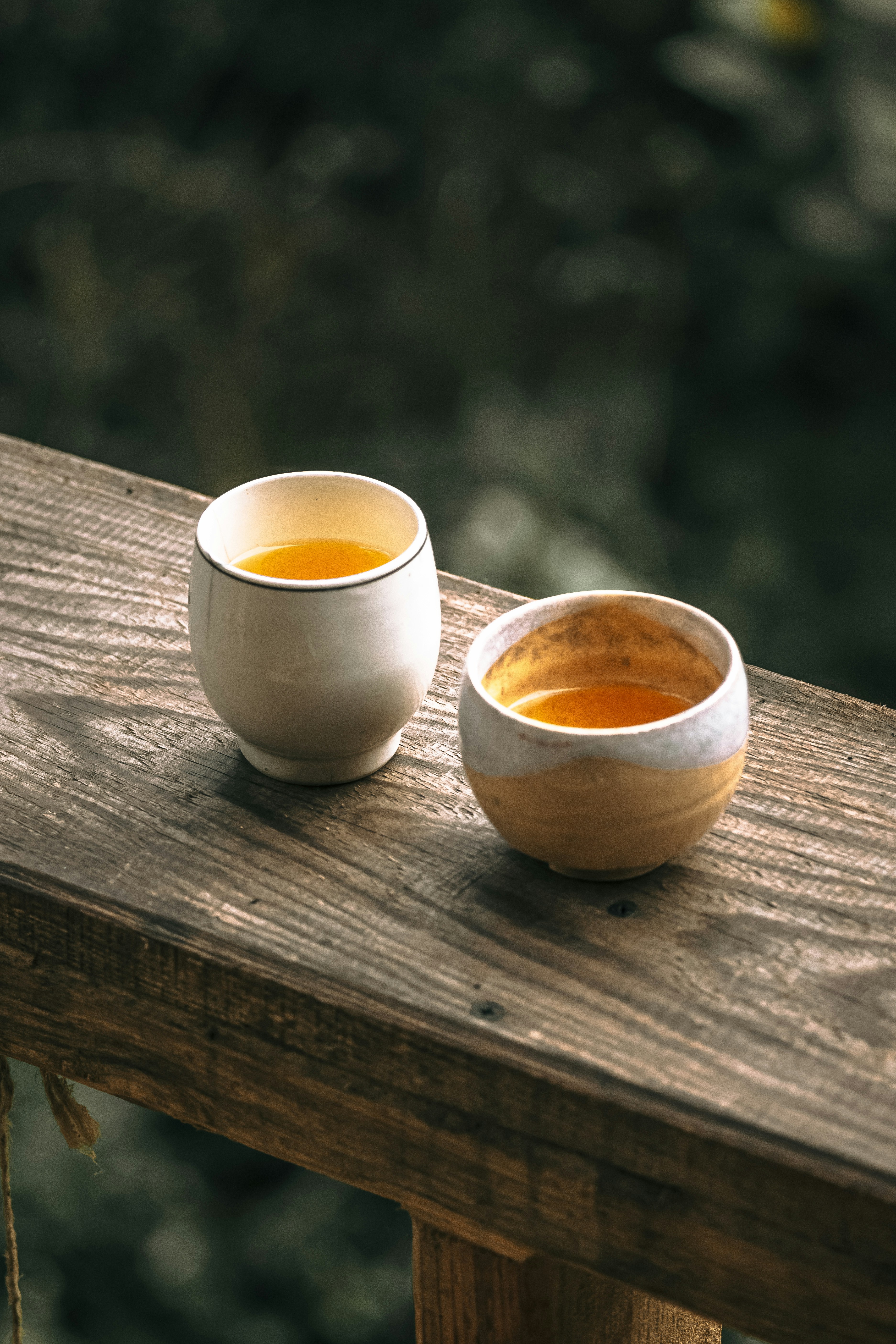 Two elegantly crafted tea cups resting on a rustic wooden surface, filled with golden tea, surrounded by a softly blurred natural backdrop.