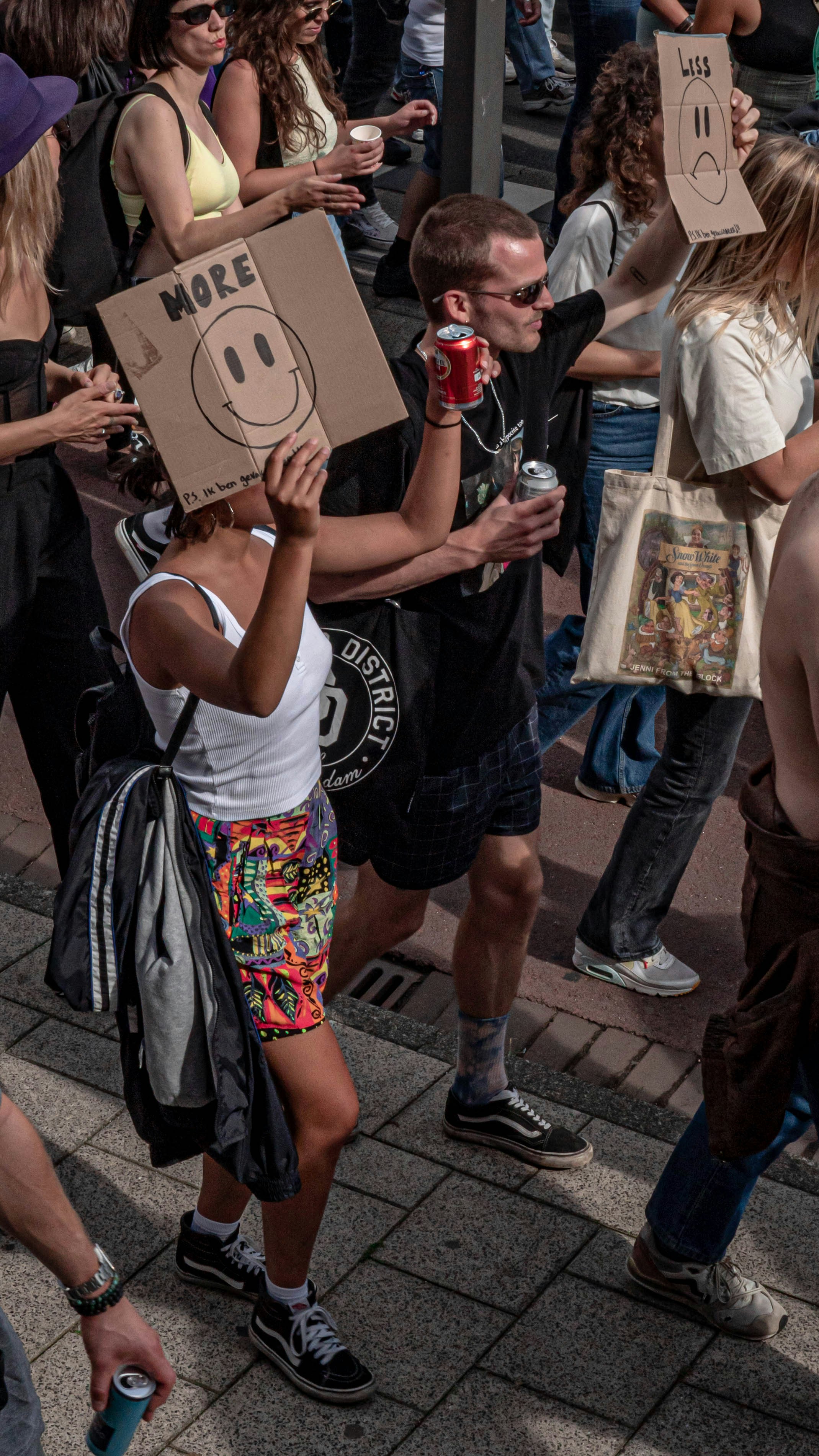 Protesters march with cardboard signs featuring a smiley, casually raising drinks and wearing casual summer attire. A tote bag and urban setting anchor the scene.