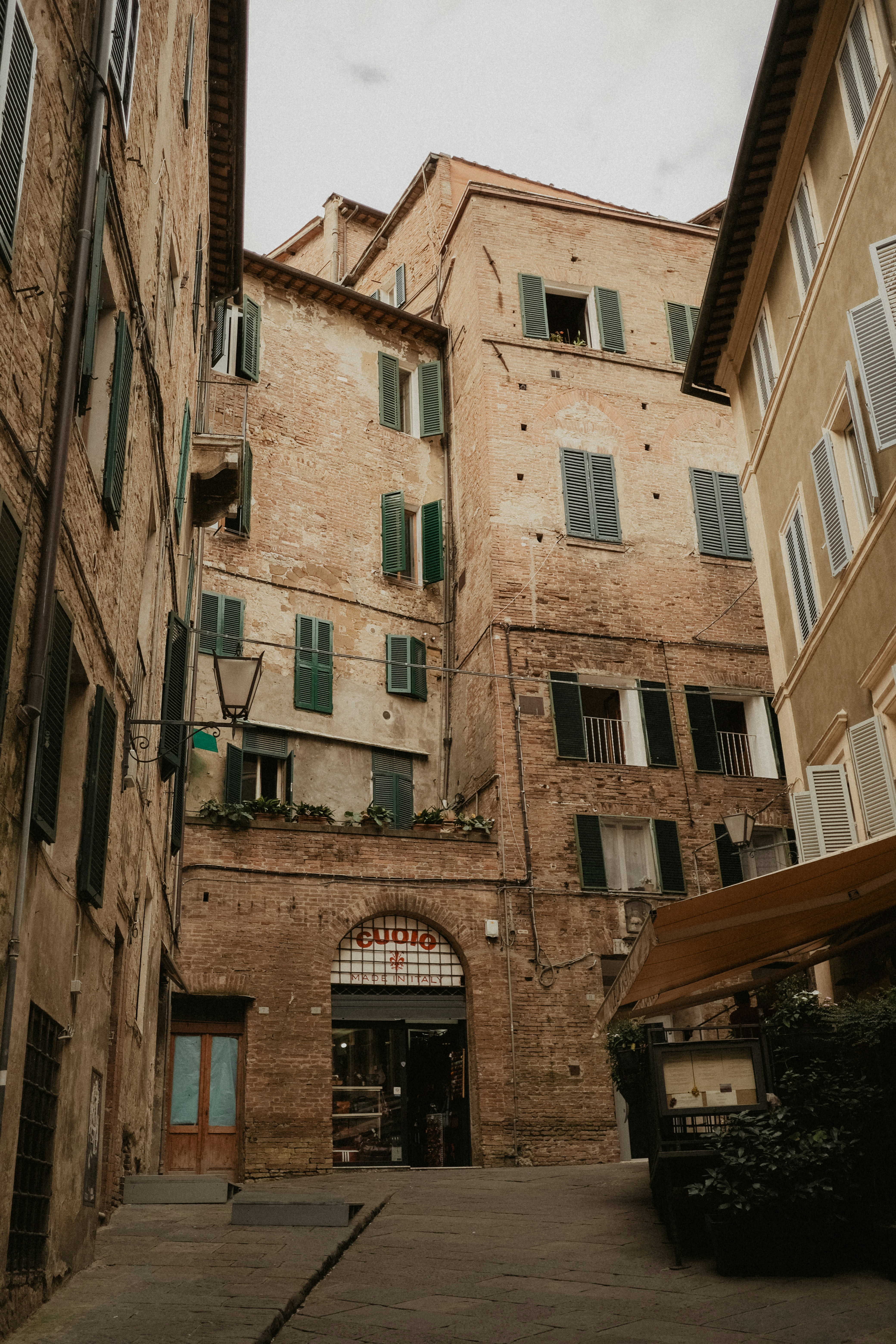 Charming alleyway showcasing rustic brick buildings adorned with green shutters, leading to a quaint shop entrance. A blend of old-world architecture and modern life.