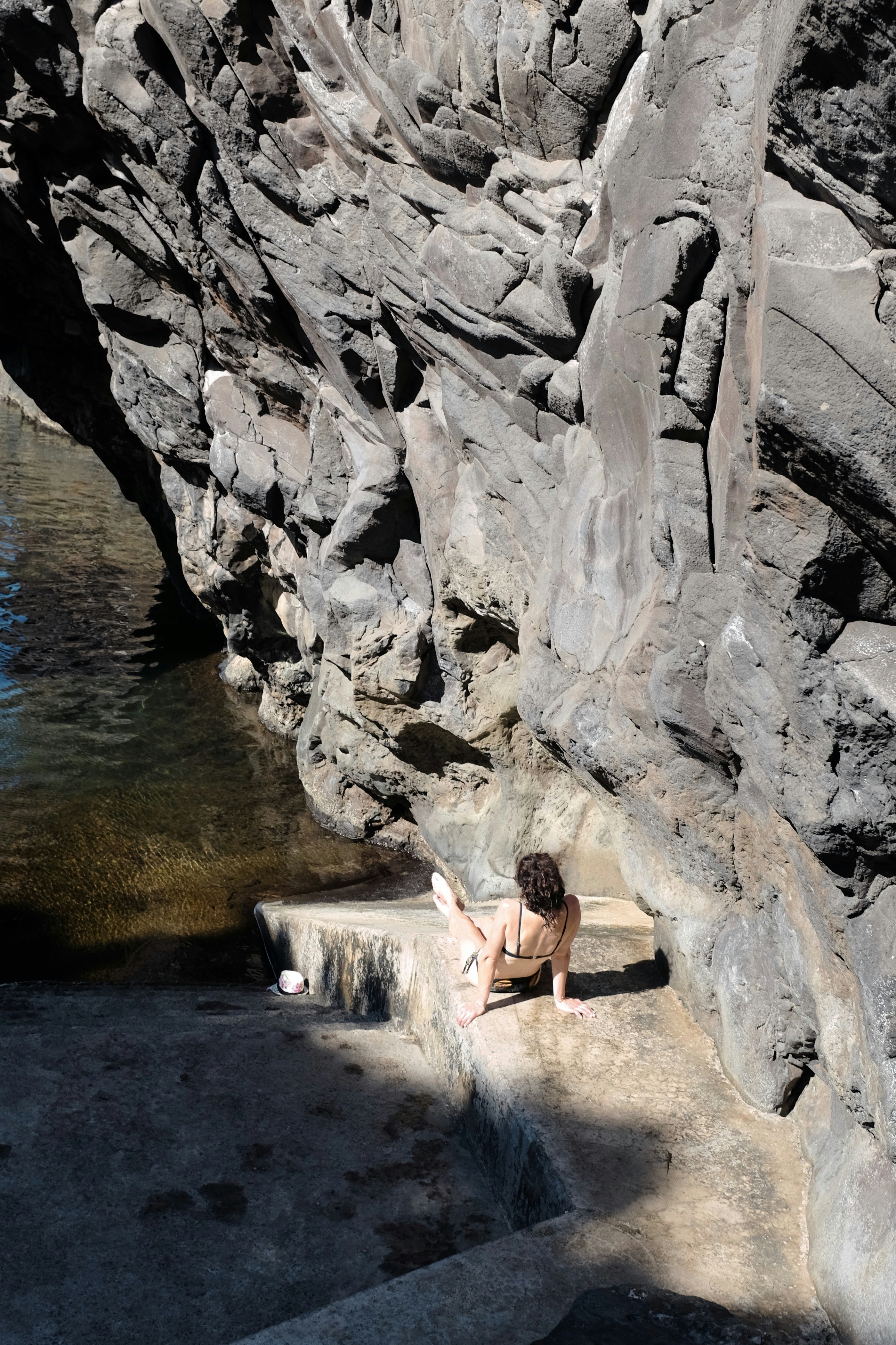 A person relaxes on stone steps beside a tranquil water pool, framed by rugged rock formations. The scene conveys a sense of peace and natural beauty.