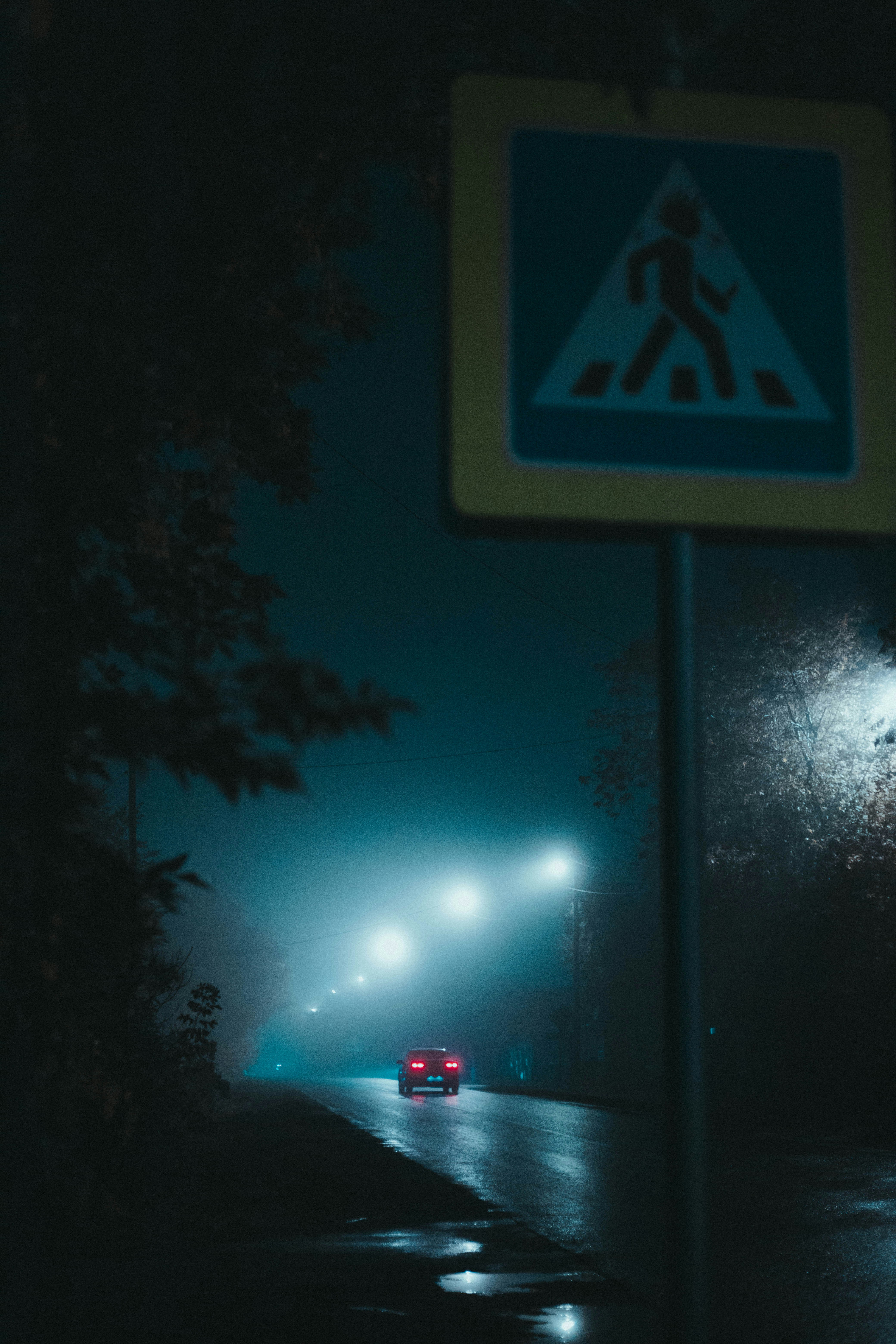 A solitary car navigates a foggy street illuminated by distant streetlights, with a roadwork sign partially obscured in the foreground.