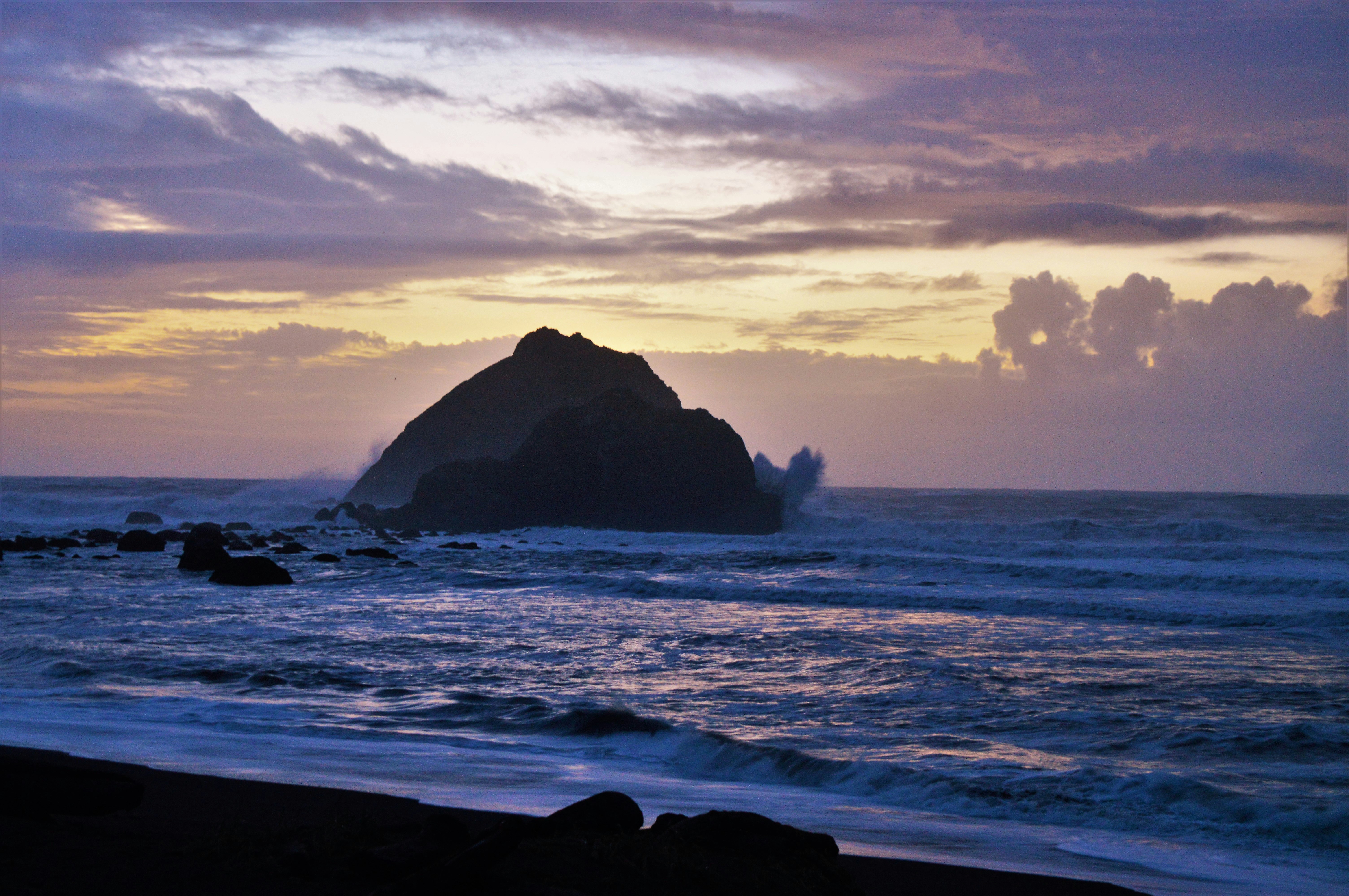 Lone rock formation rises from the ocean under a twilight sky, with waves crashing against its base. The scene captures the serene beauty of nature at dusk.