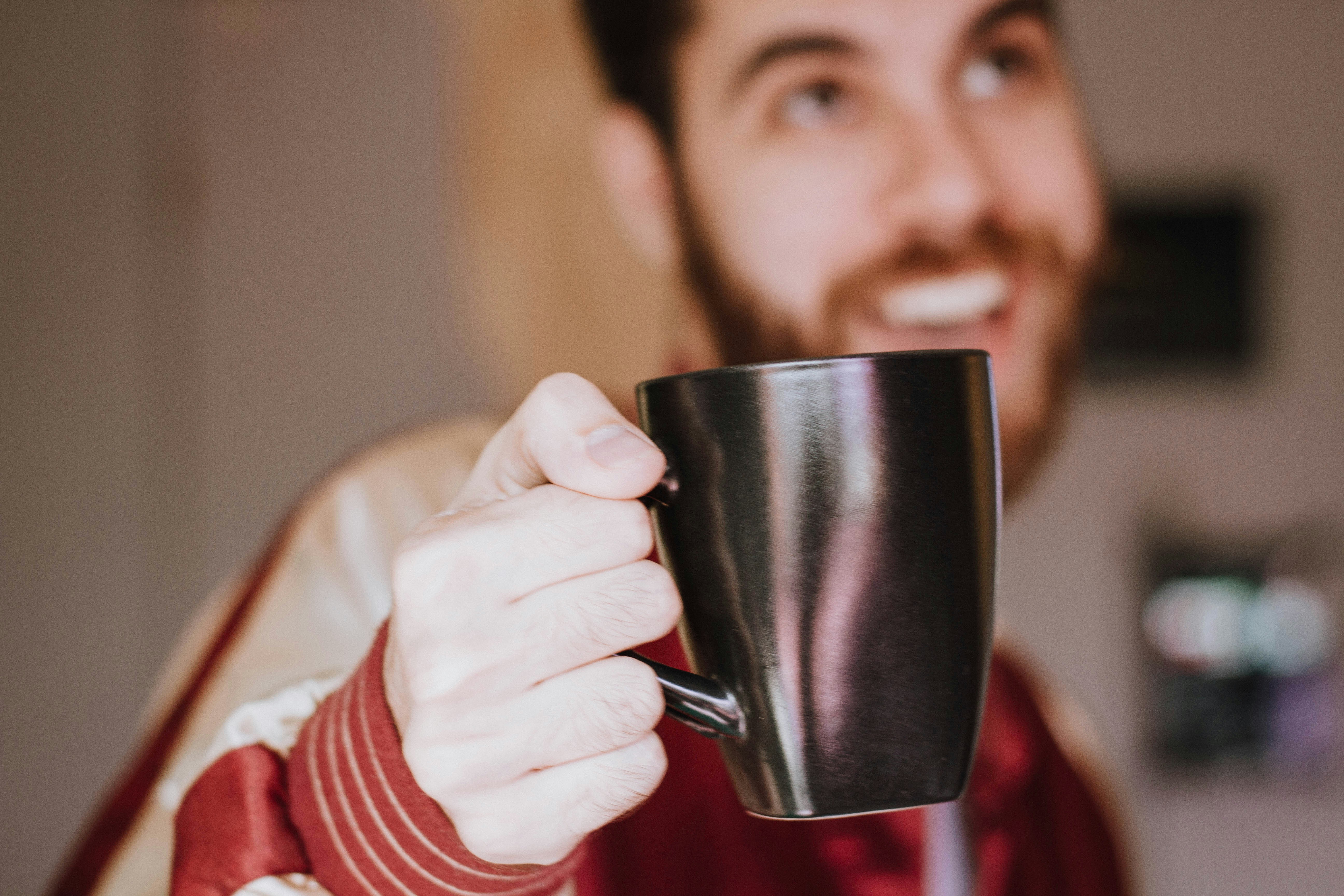 A person holding a black coffee mug, smiling in a cozy indoor setting. The soft focus background enhances the warm ambiance.