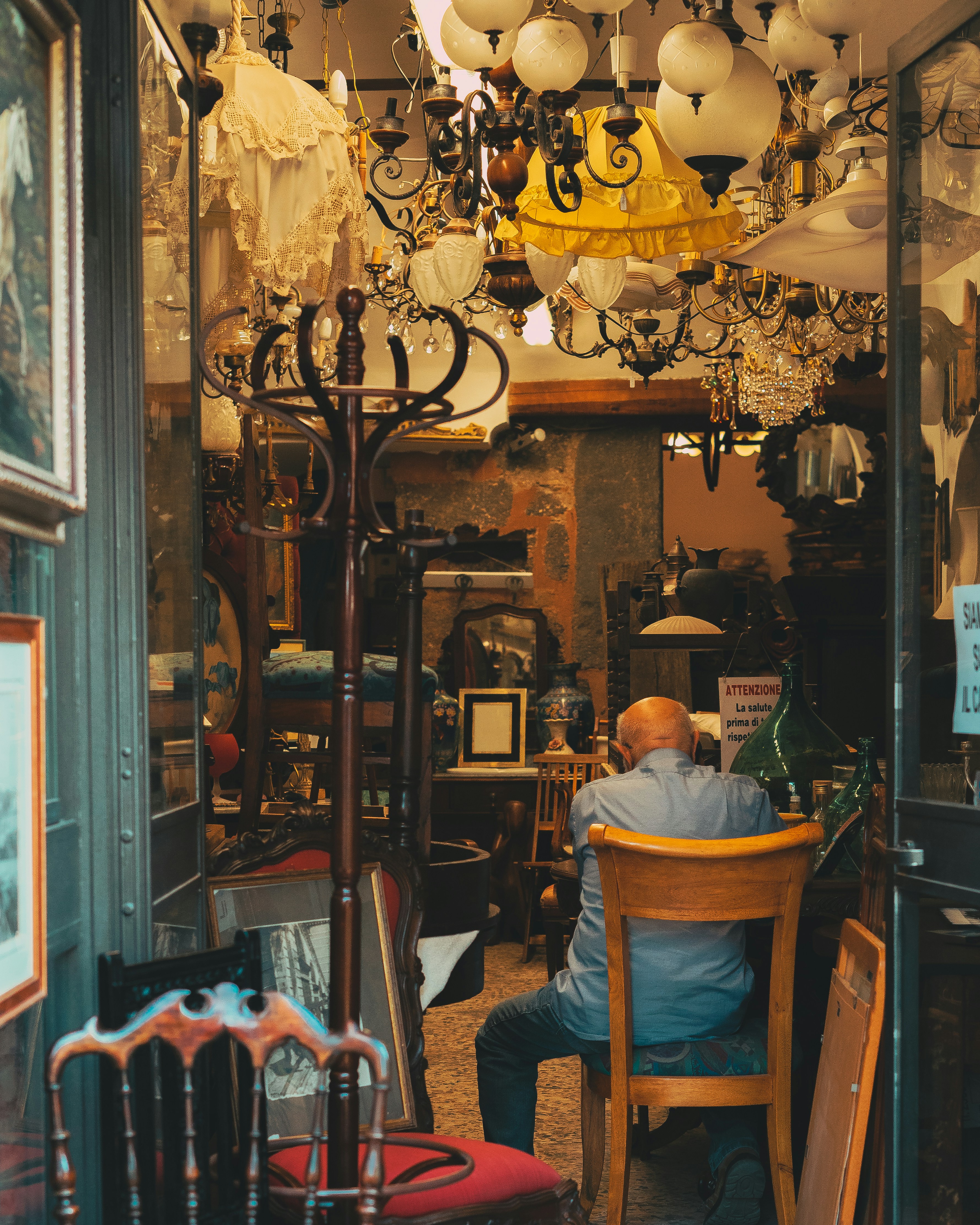 A cozy antique shop filled with vintage items and chandeliers, featuring a man seated at a wooden chair, immersed in the ambiance of nostalgia.