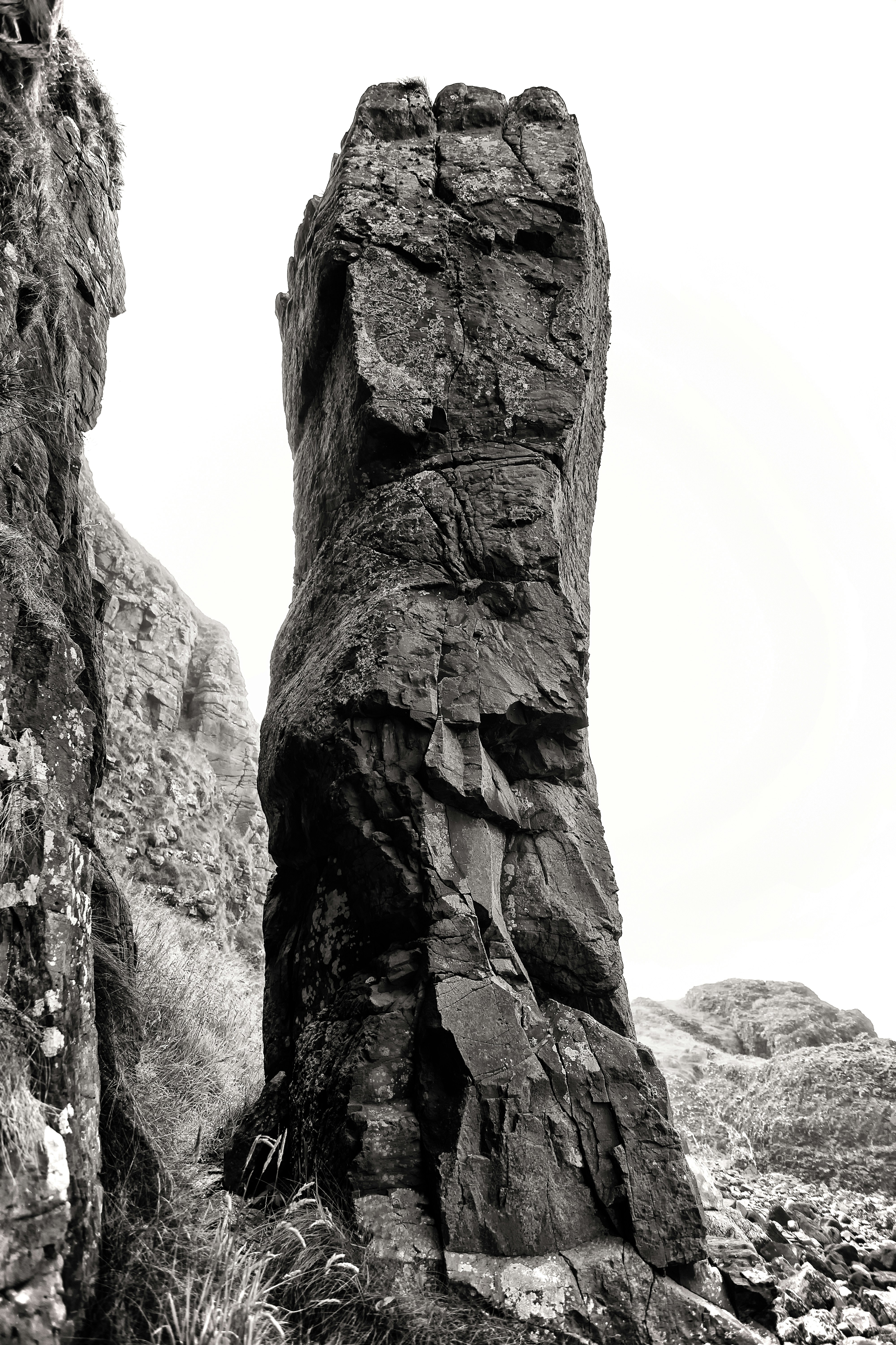 A towering rock formation stands resolute amidst rugged terrain, embodying the essence of nature's artistry. The monochrome palette accentuates the textures and contours of the stone.