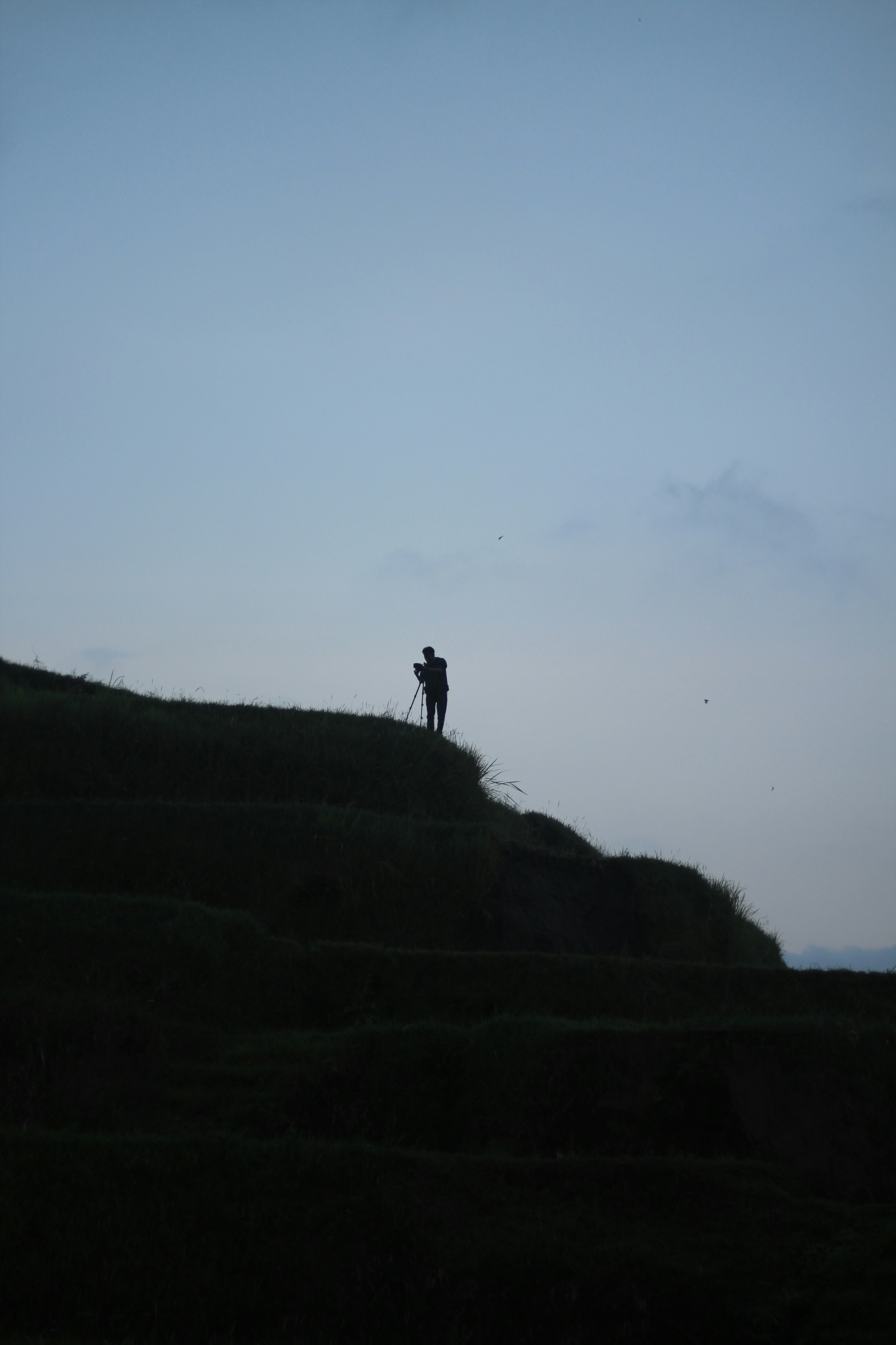 Silhouette of a photographer standing on a hillside, capturing the serene twilight sky. The layered terrain adds depth to the composition.